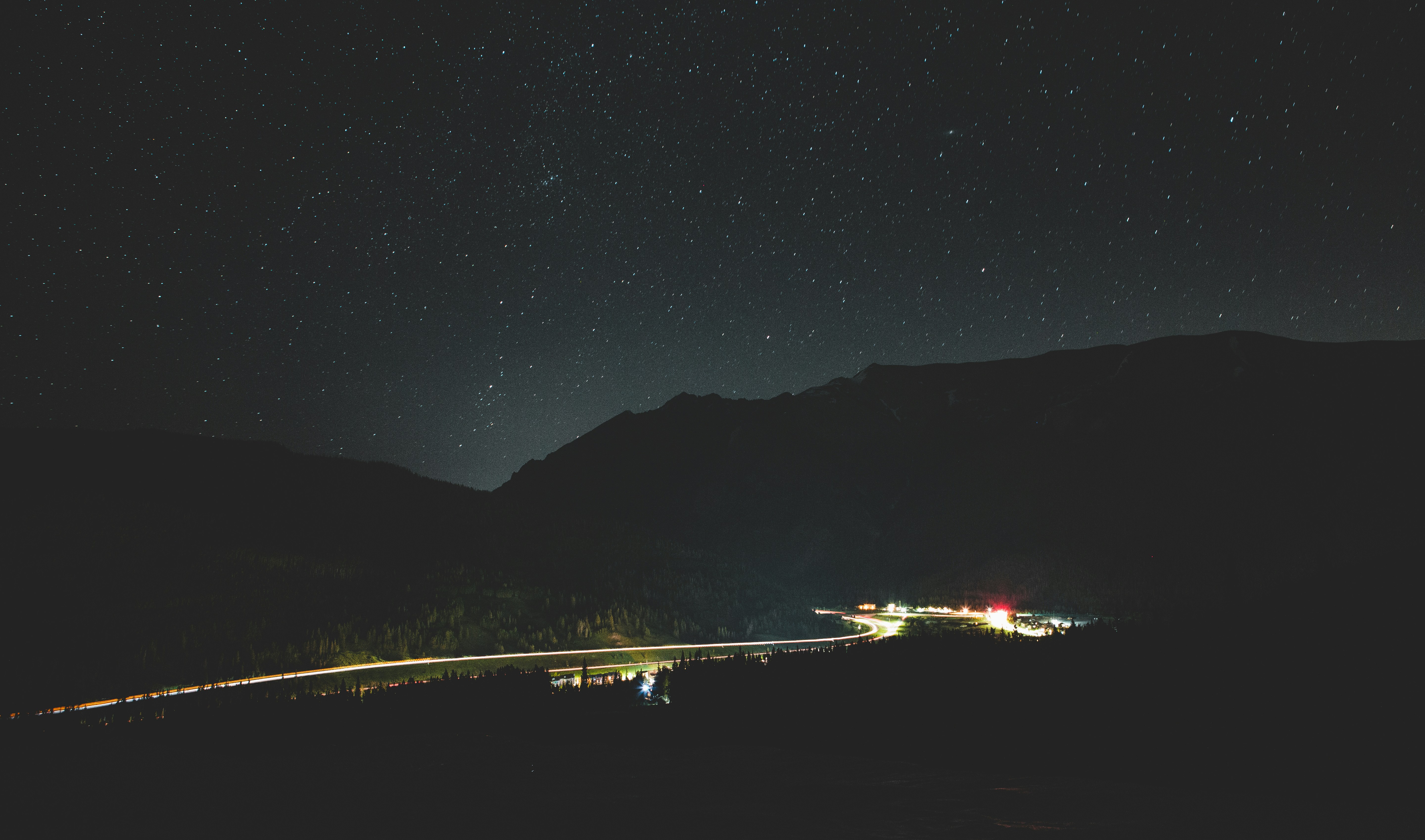 Starry night sky over a winding mountain road illuminated by vehicle lights.