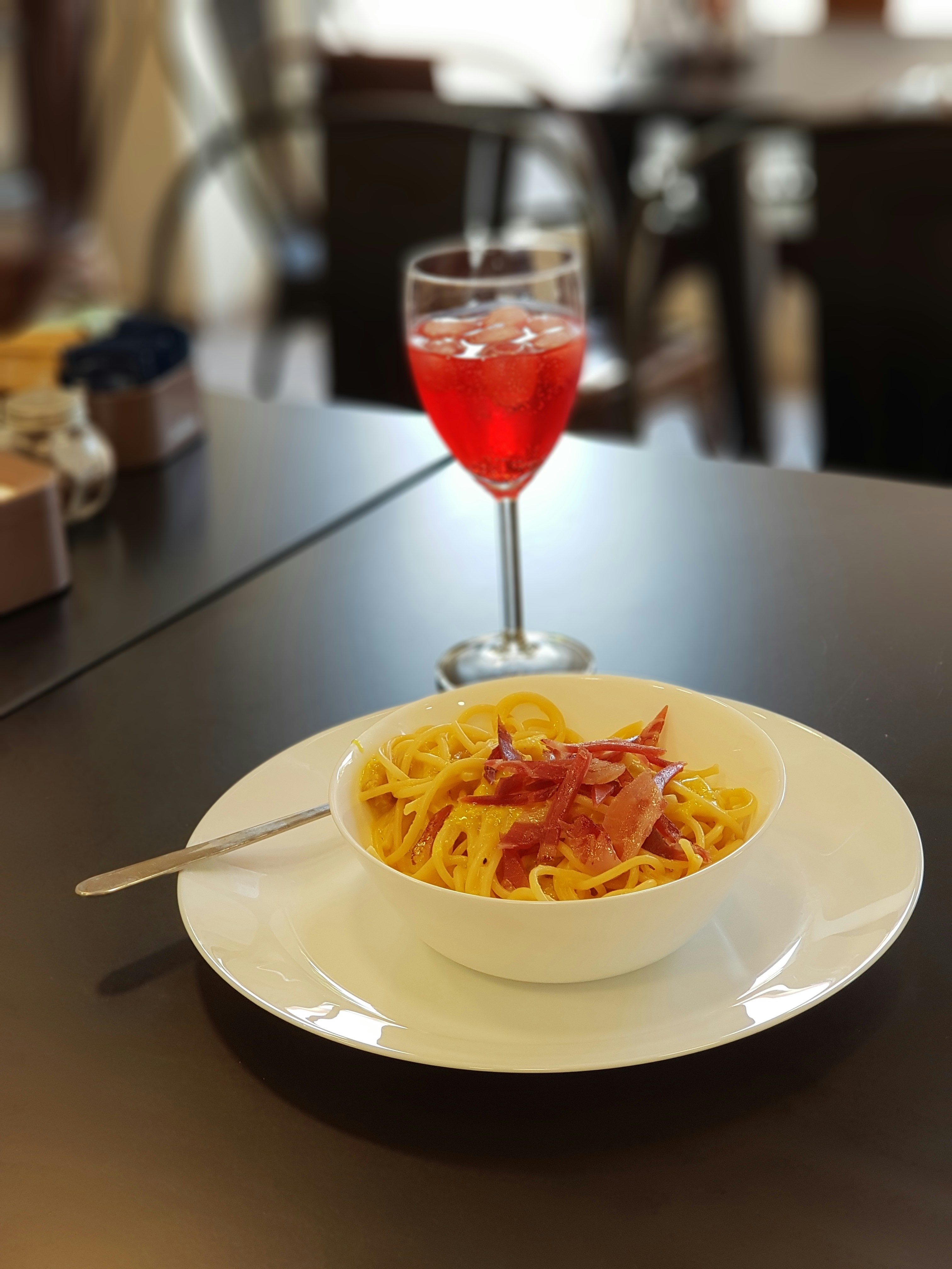 A bowl of fettuccine topped with crispy bacon sits elegantly on a table, accompanied by a vibrant red drink in a stemmed glass.