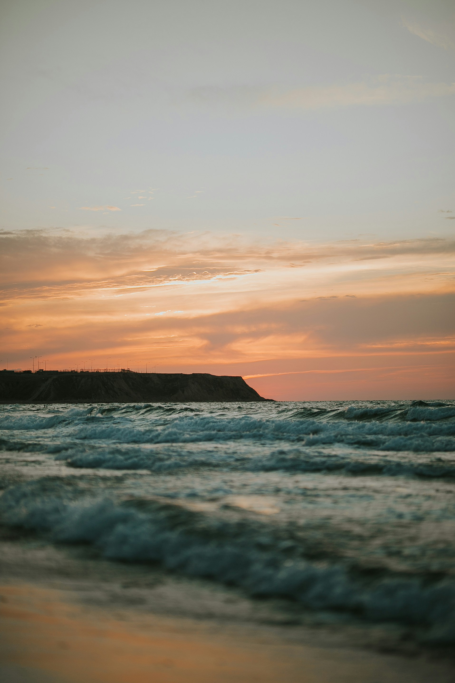 Gentle waves lapping at the shore under a pastel sky as the sun sets behind a distant cliff.