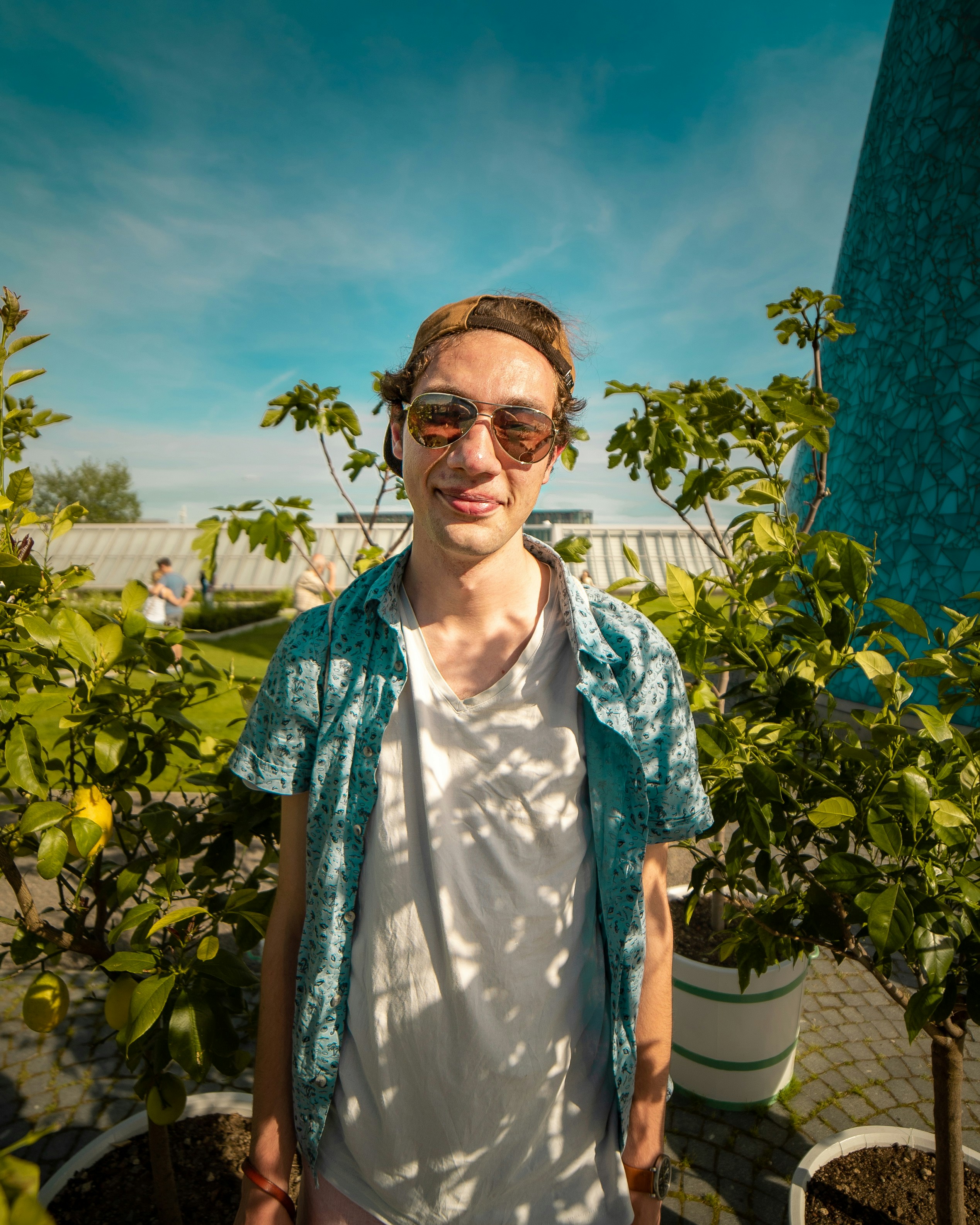Young man in sunglasses stands amidst vibrant greenery, enjoying a sunny day. The backdrop features a unique architectural element.