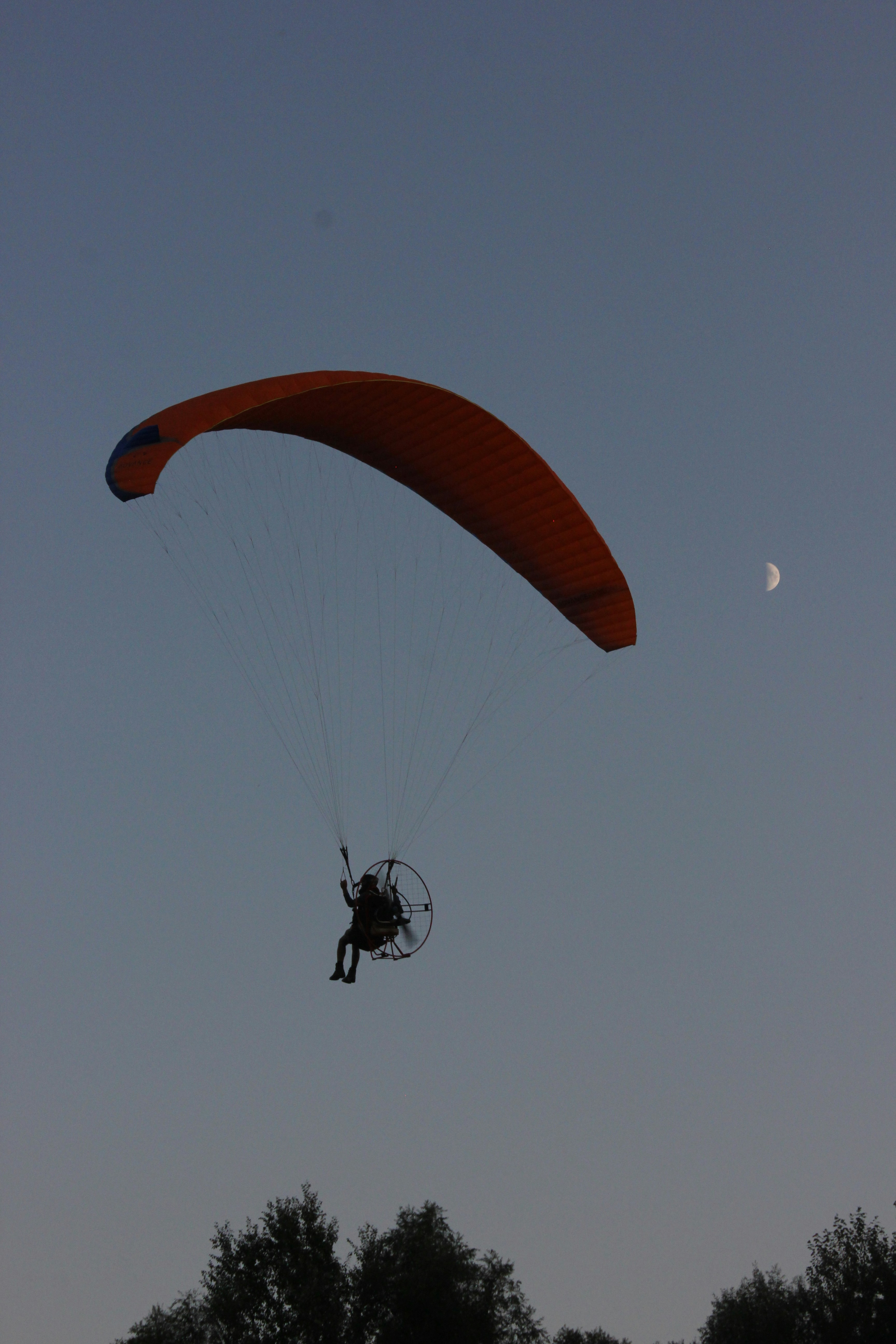 A paramotorist gracefully glides through the twilight sky beneath a crescent moon, silhouetted against the fading light. The vibrant orange canopy contrasts with the serene blue backdrop.
