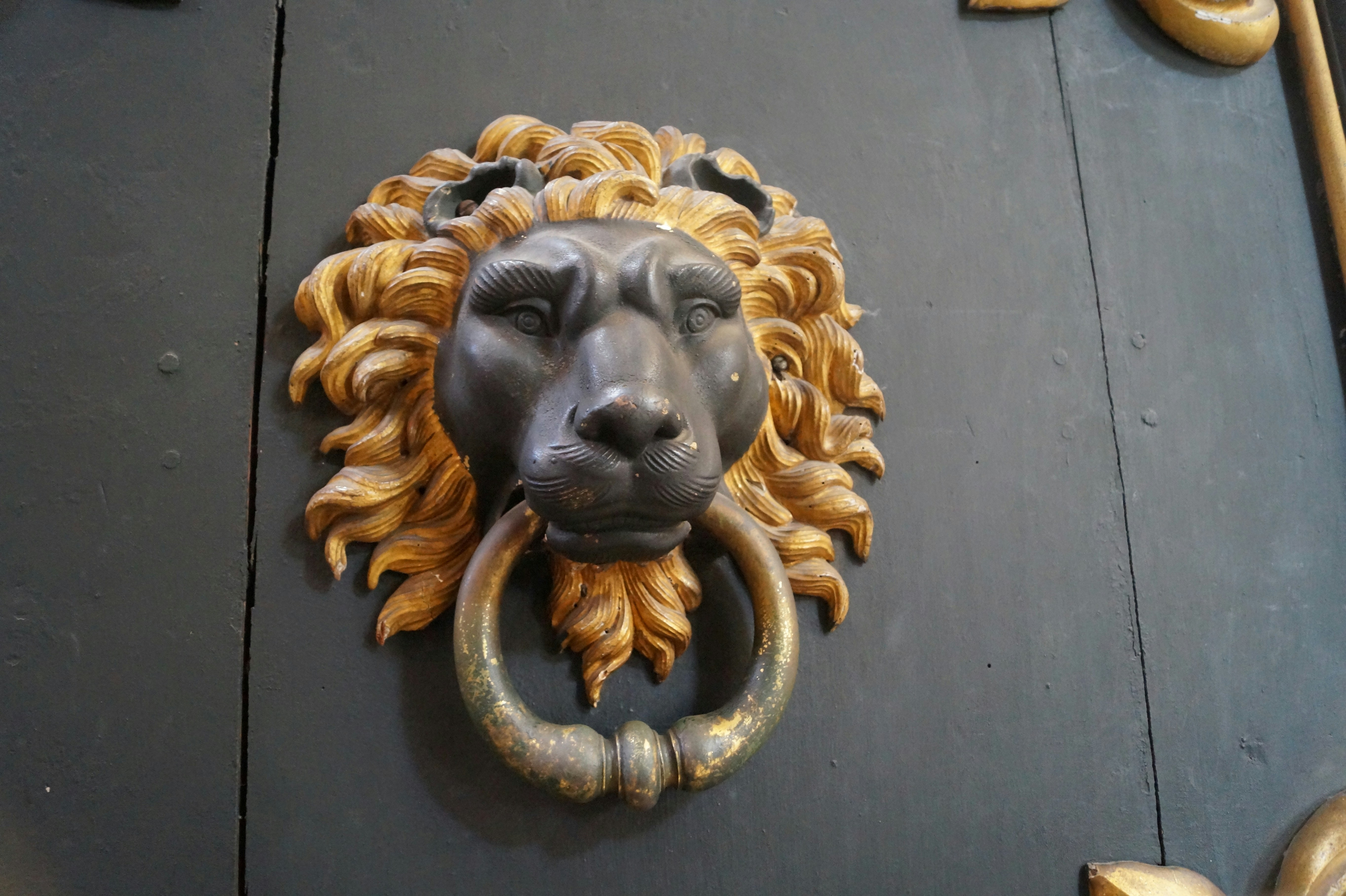 Intricately designed lion head door knocker featuring golden mane and detailed facial features, mounted on a dark wooden door.