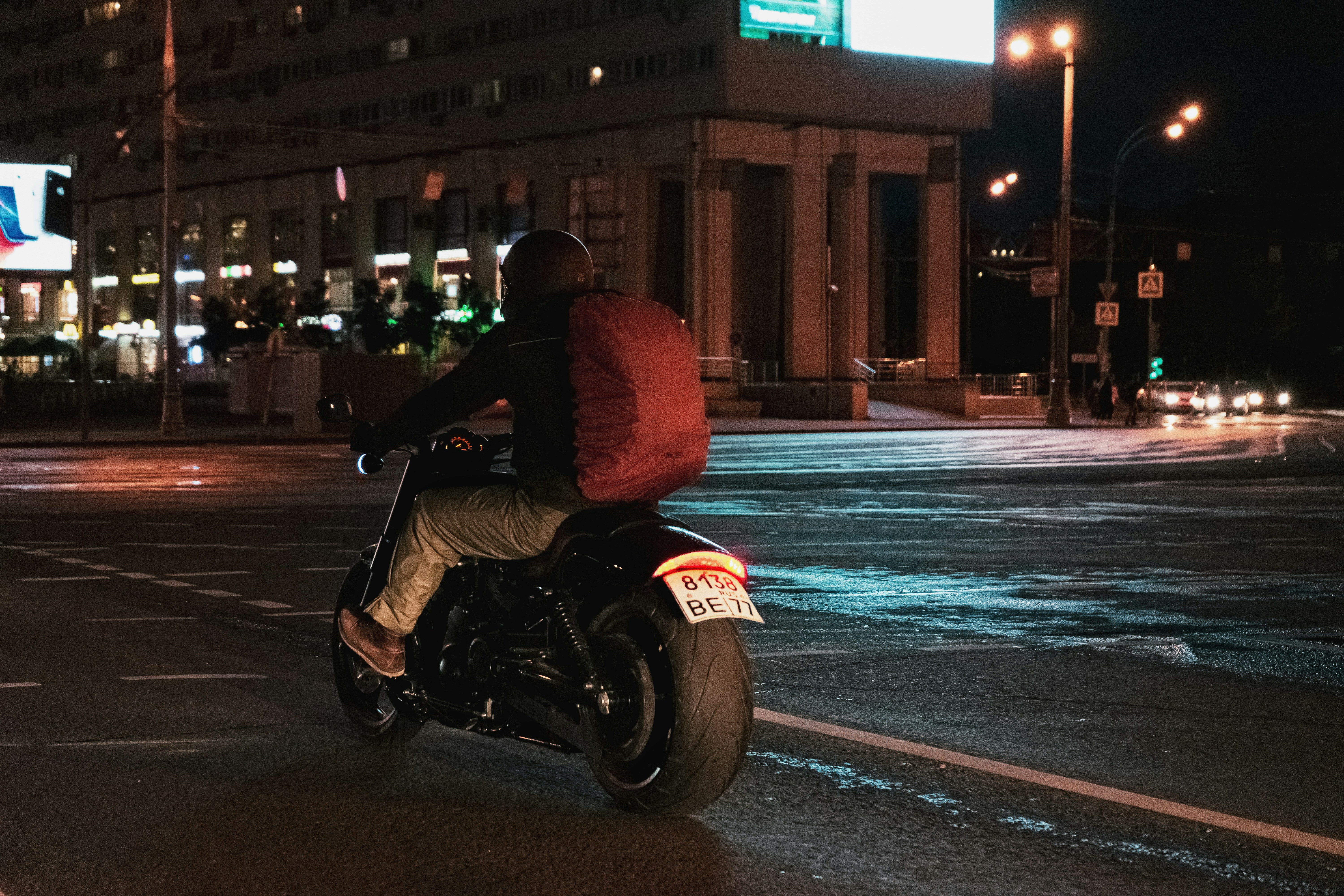 Motorcyclist with a red backpack navigating a city intersection at night, illuminated by streetlights and neon signs.