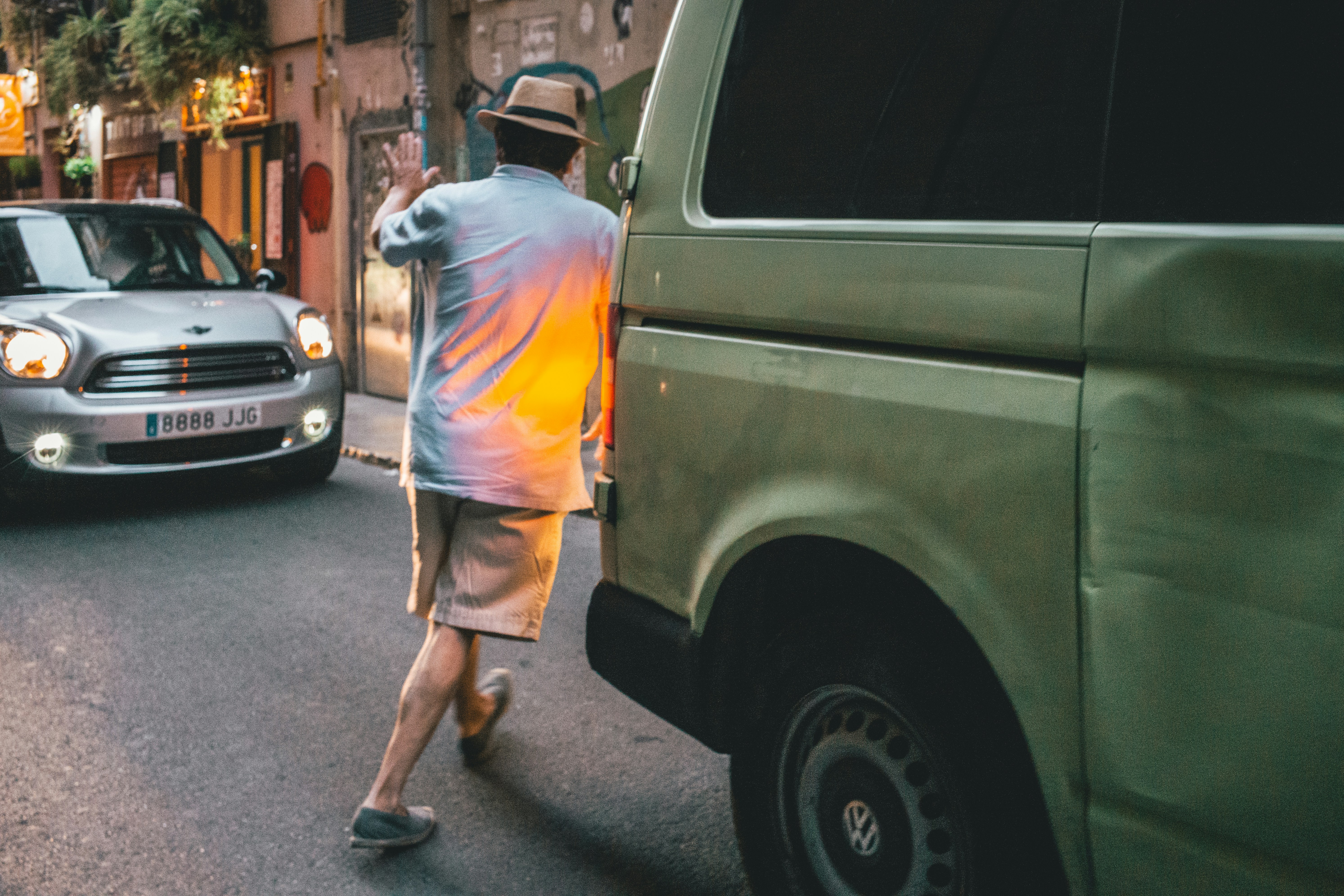 Street photograph captures a man in a hat and light blue shirt moving past a green van on a narrow urban street at dusk.