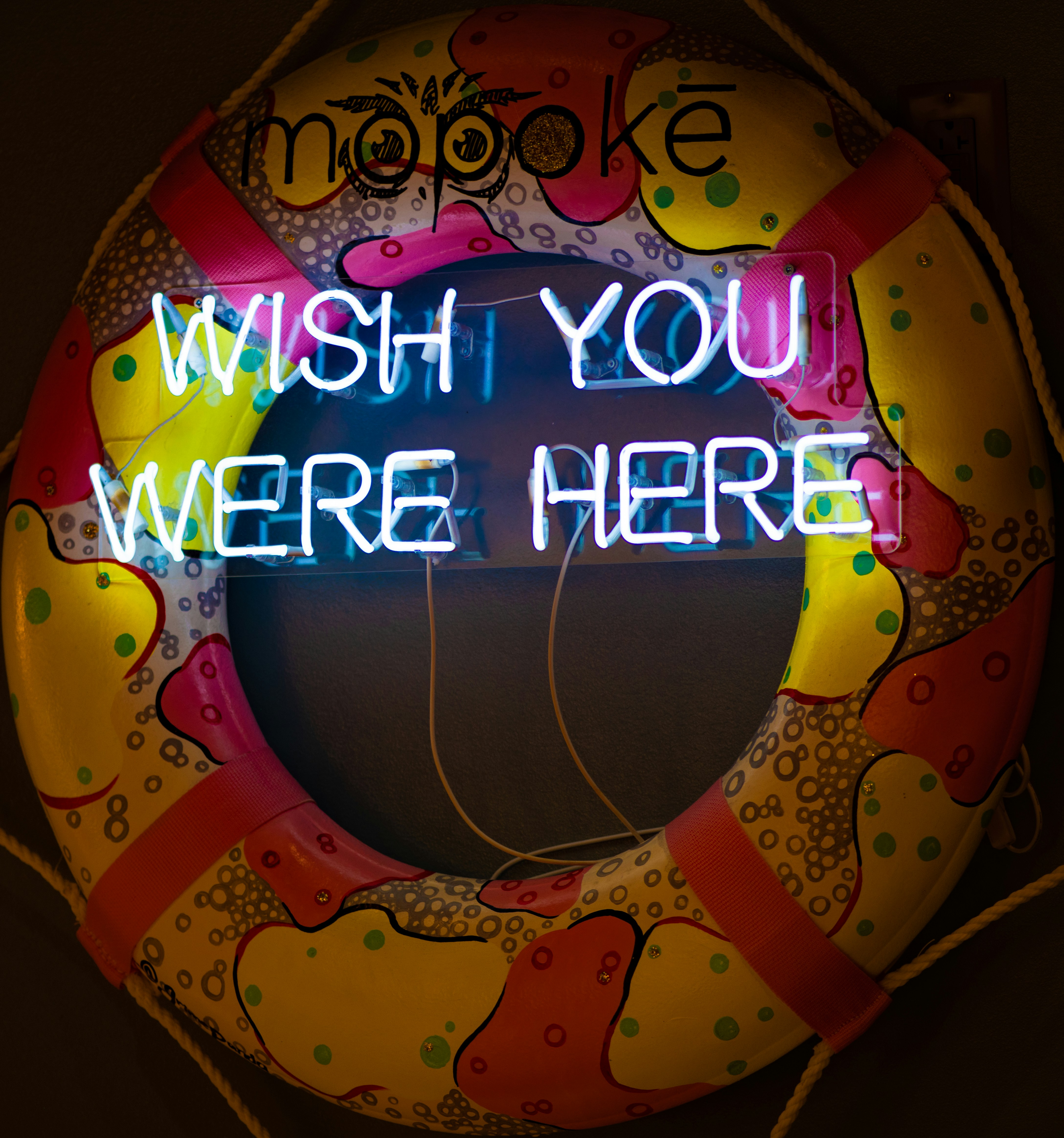 A colorful lifebuoy encircles a glowing neon sign that reads 'WISH YOU WERE HERE.' The vibrant design adds a playful touch to the scene.