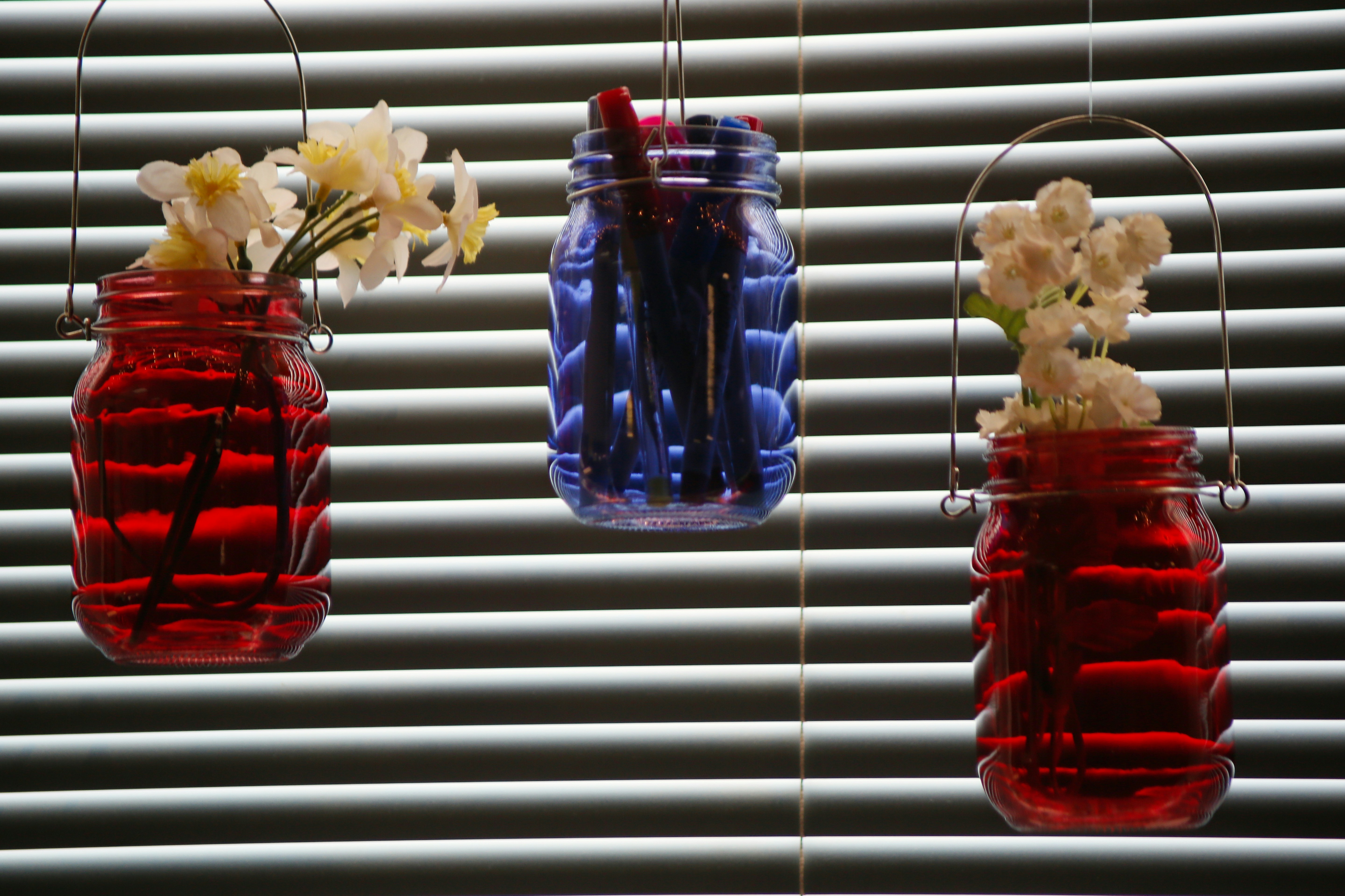 Three hanging jars filled with flowers and pens, set against a backdrop of horizontal blinds. The interplay of red and blue adds a vibrant touch.