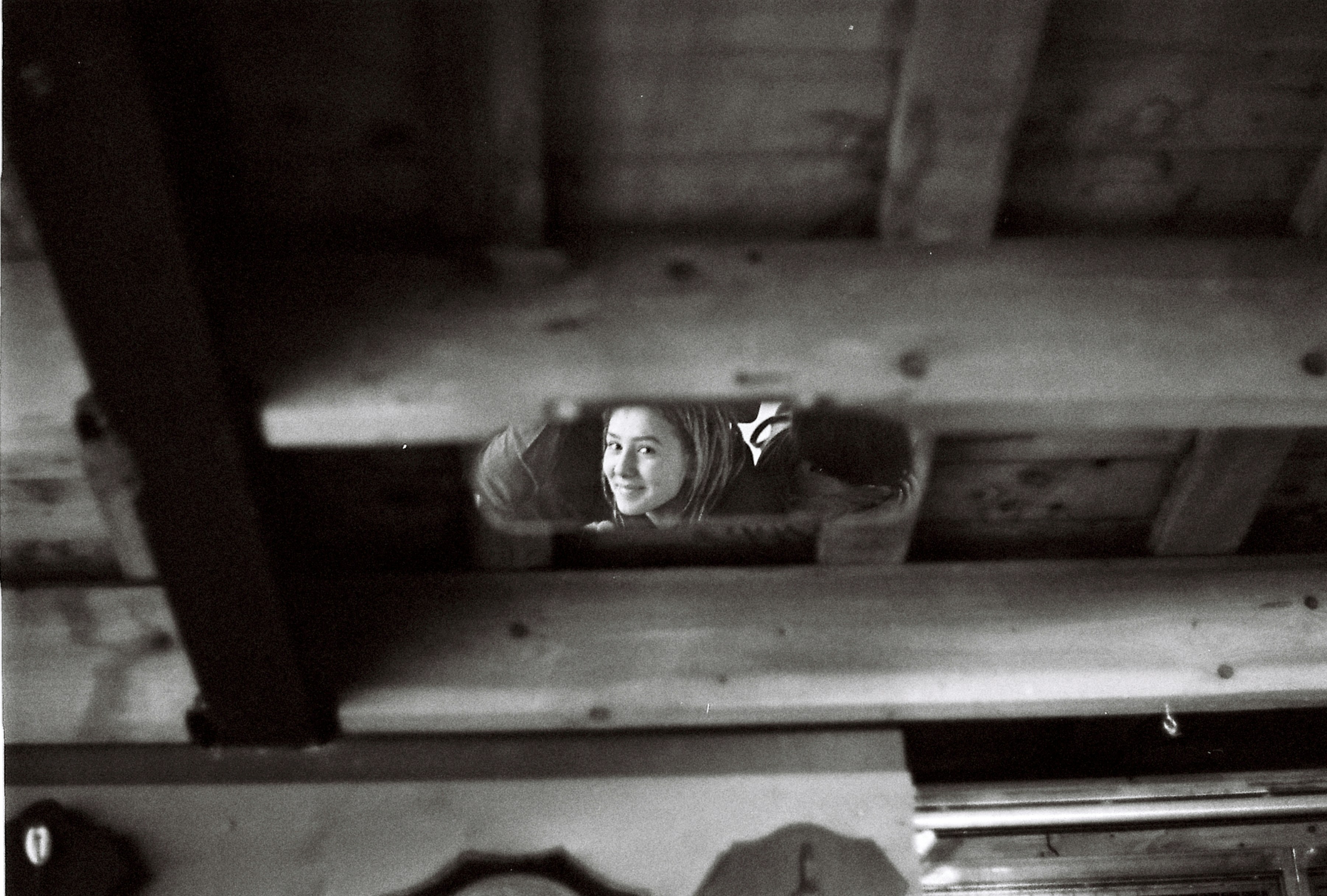 A smiling face peeks through a wooden beam, framed by rustic architecture. The monochrome tones enhance the warmth of the moment.