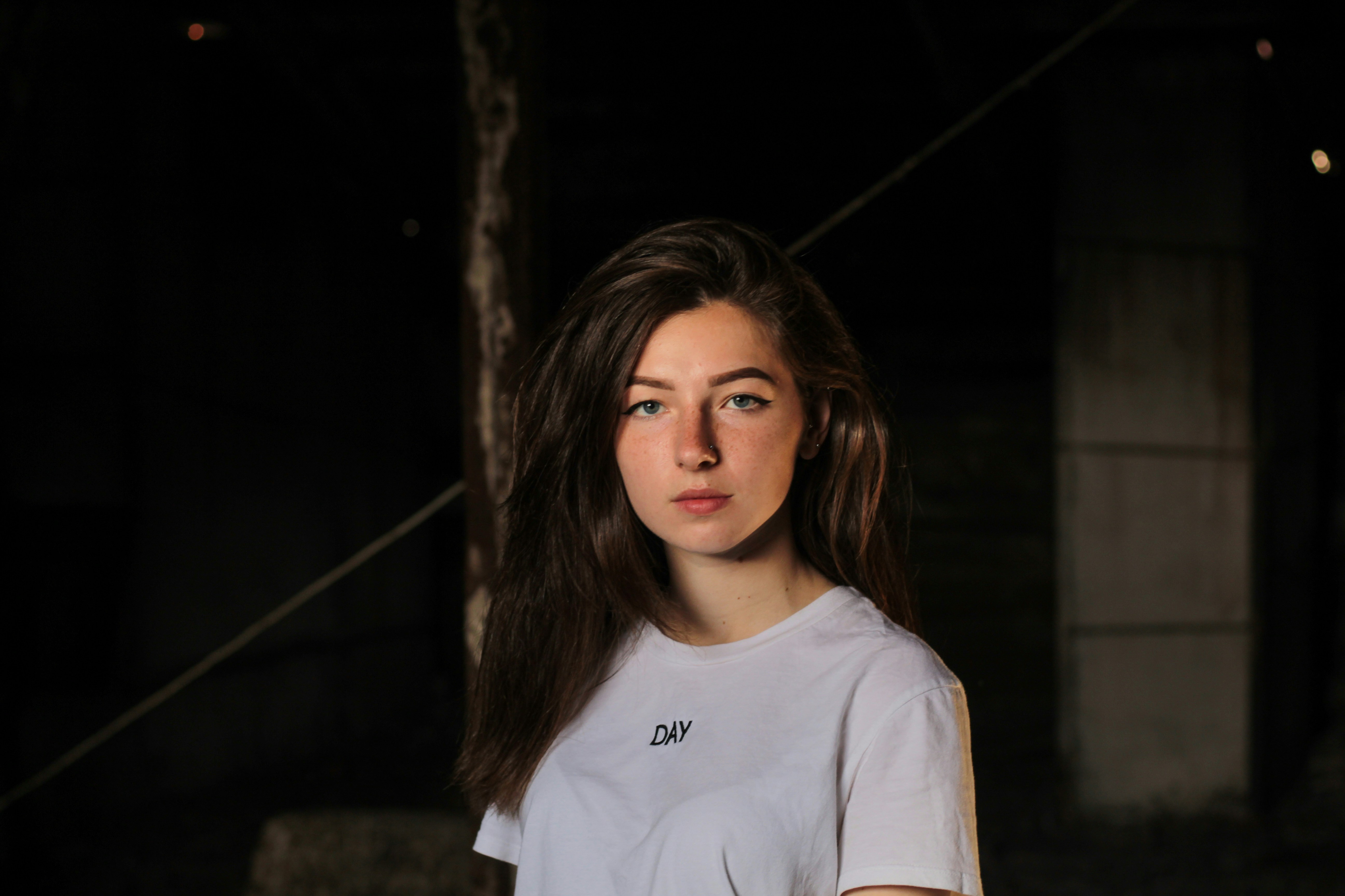 Young woman with long hair wearing a white t-shirt stands confidently in a dimly lit industrial space.
