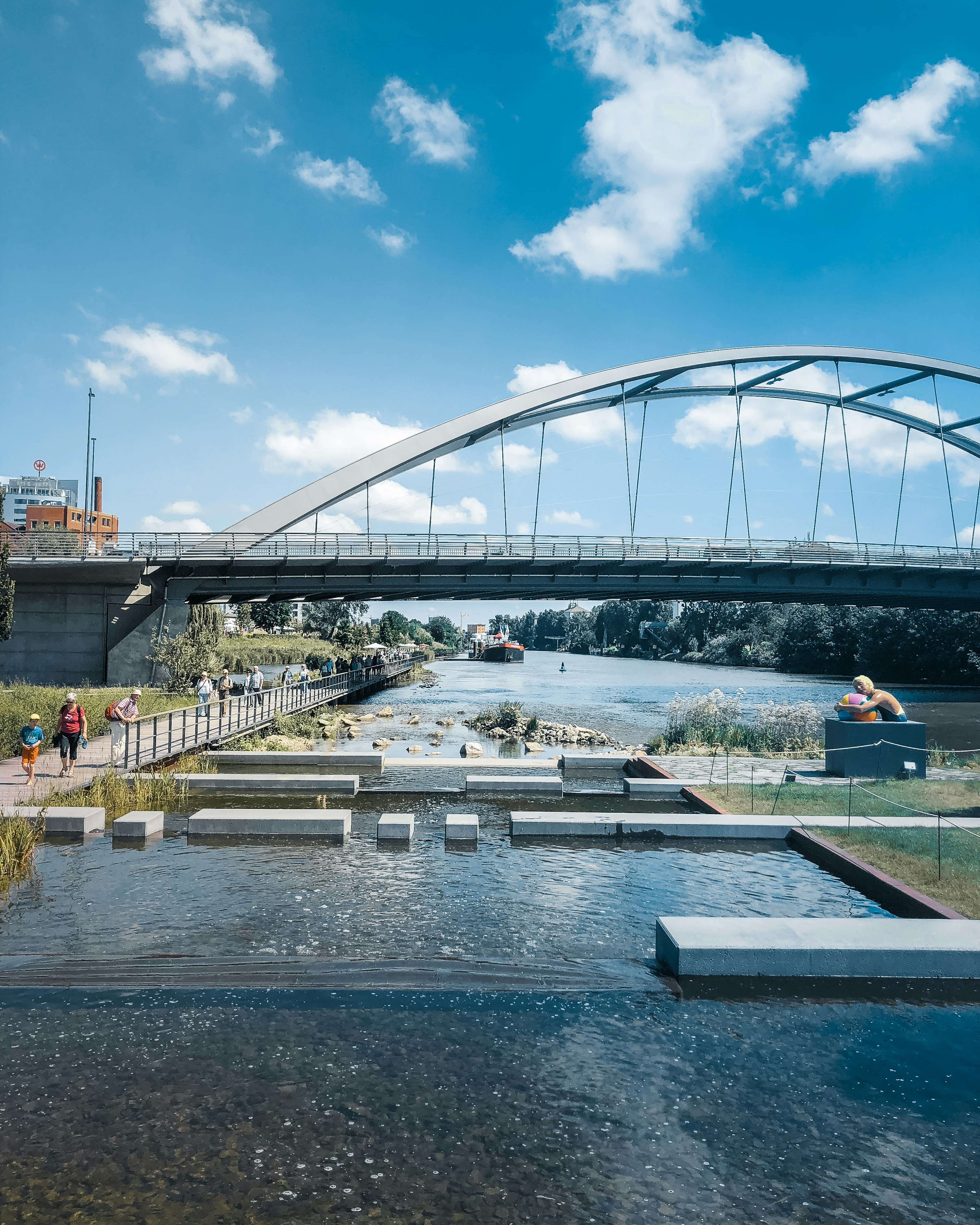A modern arch bridge spans a serene river, with pedestrians enjoying a scenic walkway alongside flowing water and greenery.