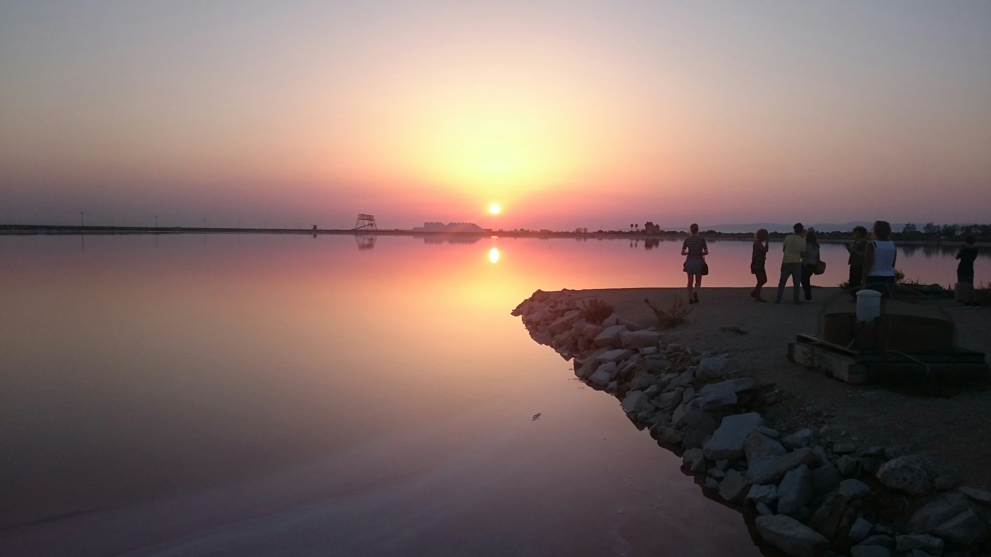 Sunset casting a warm glow over a calm body of water with silhouettes of people along the shore. The tranquil scene invites reflection.