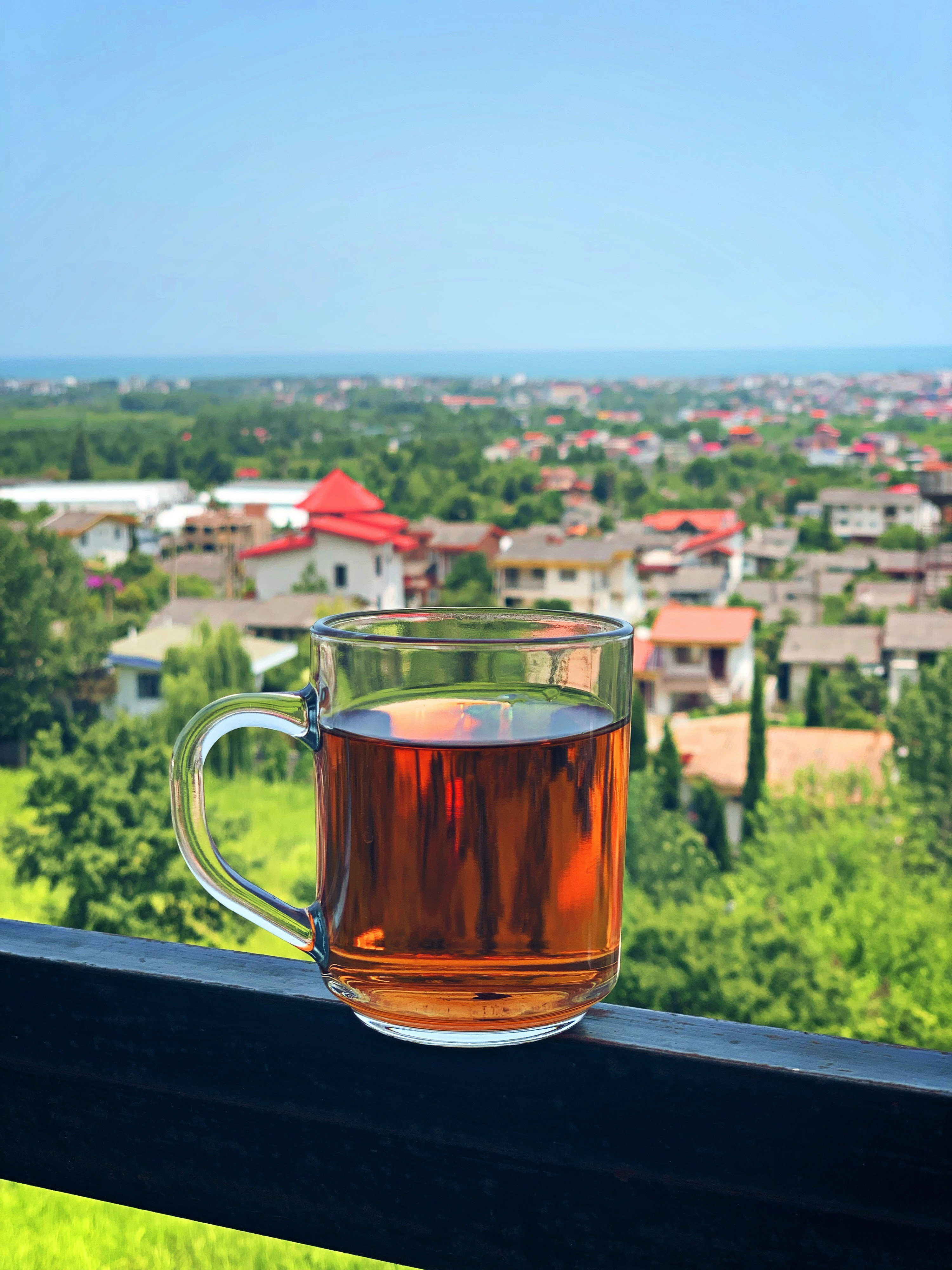 Clear glass mug of tea on a balcony railing overlooking a vibrant townscape and distant horizon.