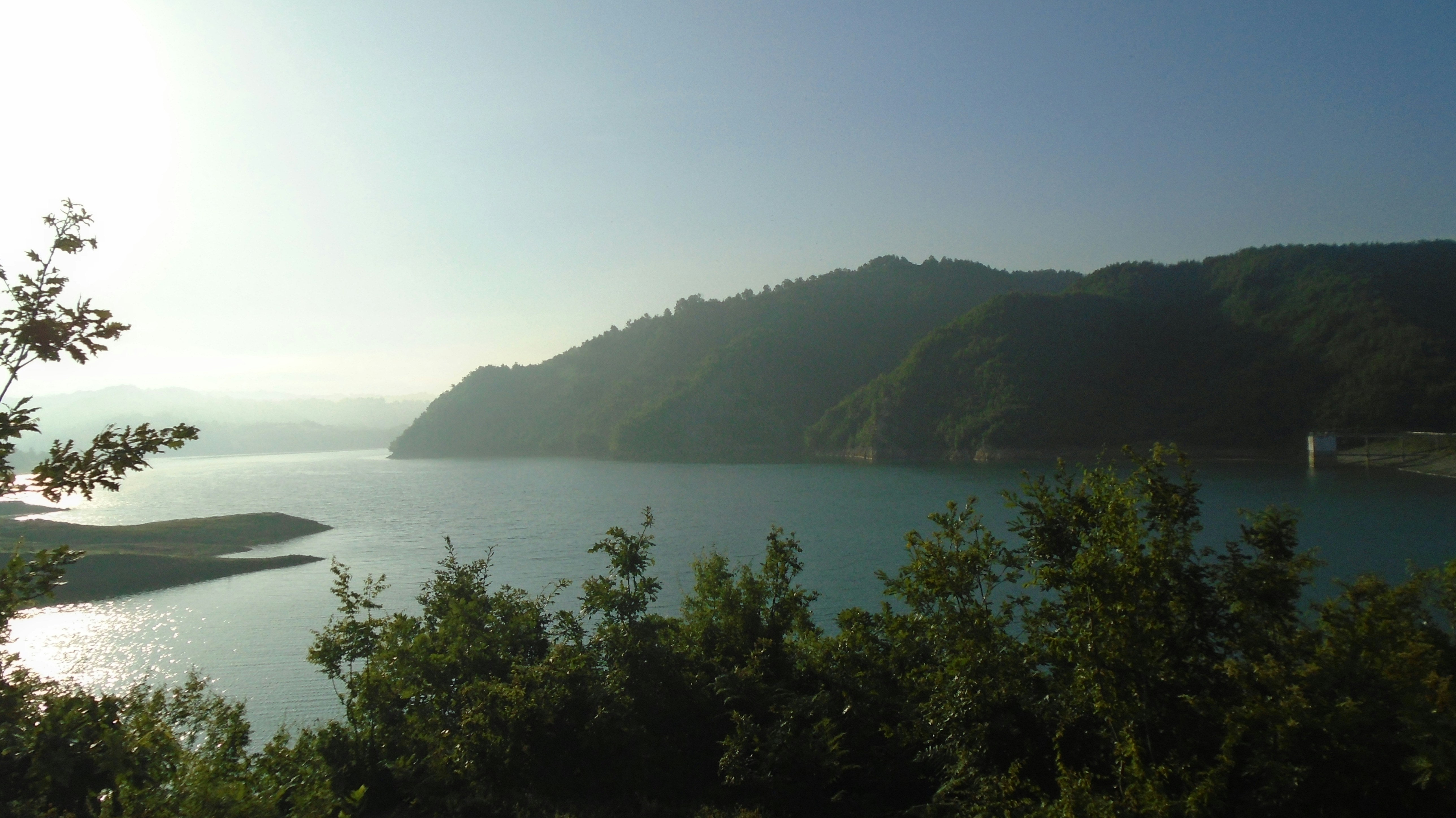Tranquil lake scene framed by lush greenery and distant hills, capturing the essence of a peaceful morning. The soft light enhances the calm waters.