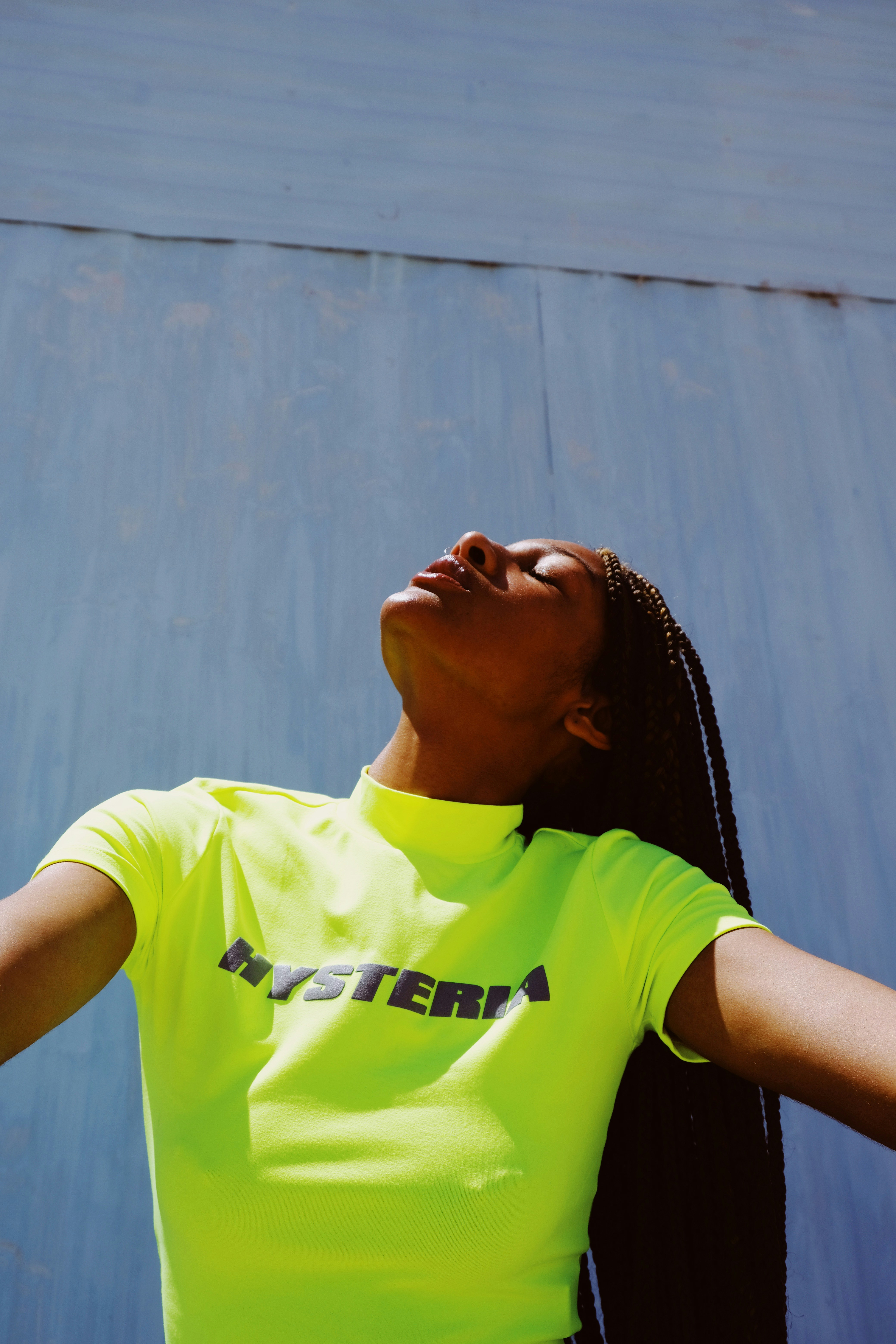 A woman in a vibrant neon shirt tilts her head back, basking in sunlight against a blue backdrop.