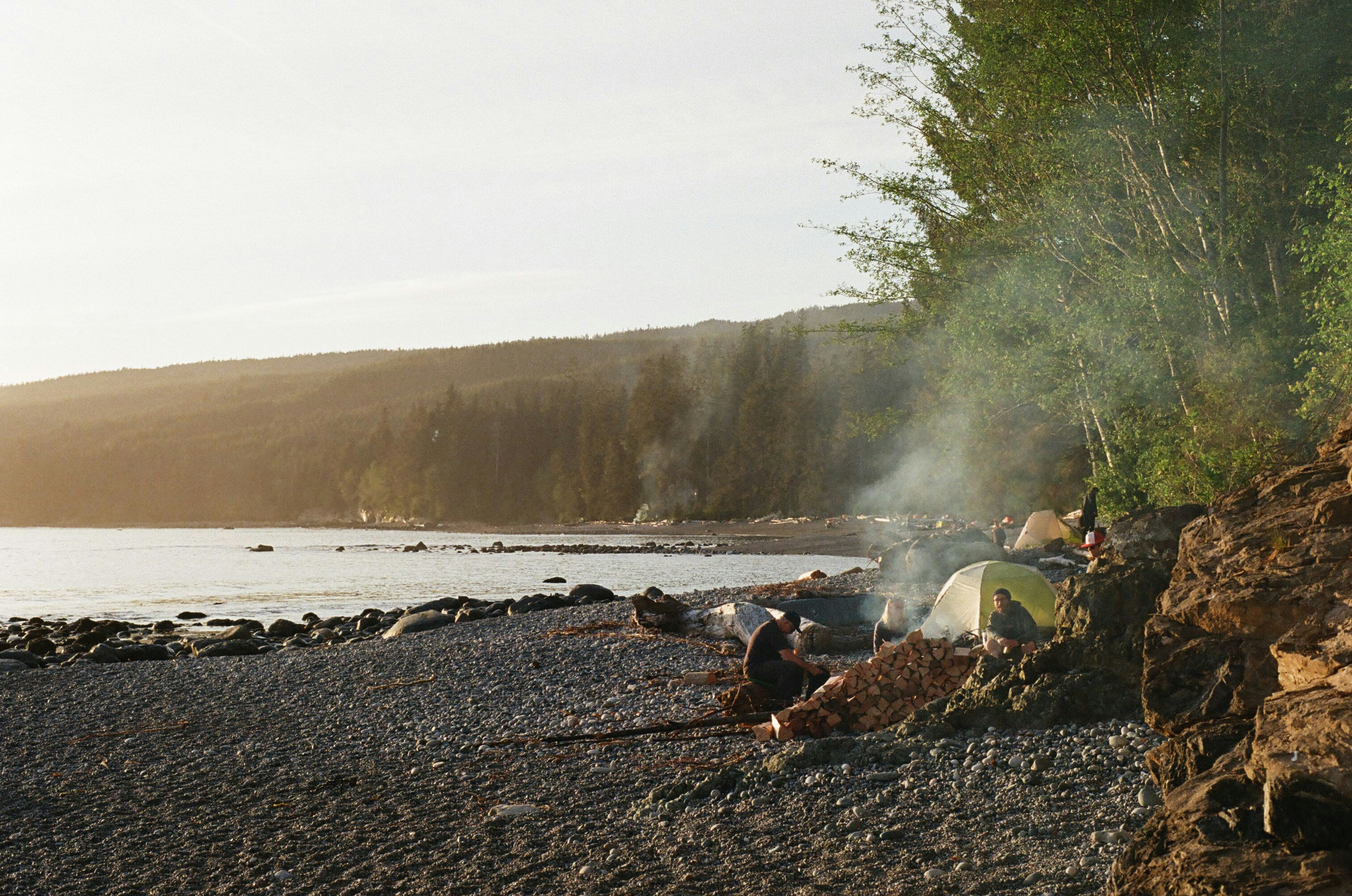 Tents and smoke by a pebbled shoreline at sunset