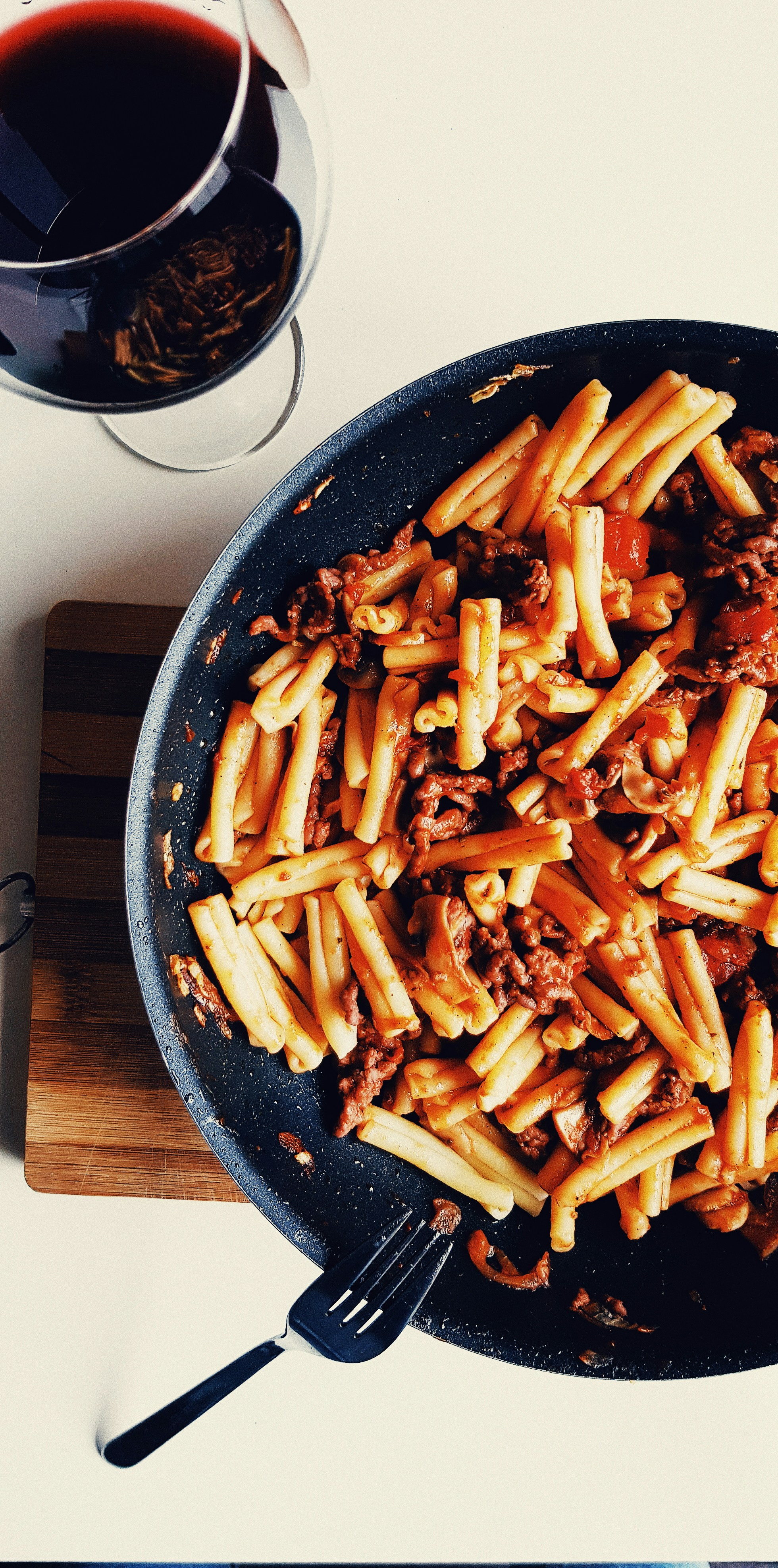 Delicious pasta dish with ground meat and tomatoes served in a frying pan, accompanied by a glass of red wine. The setting suggests a cozy dining experience.