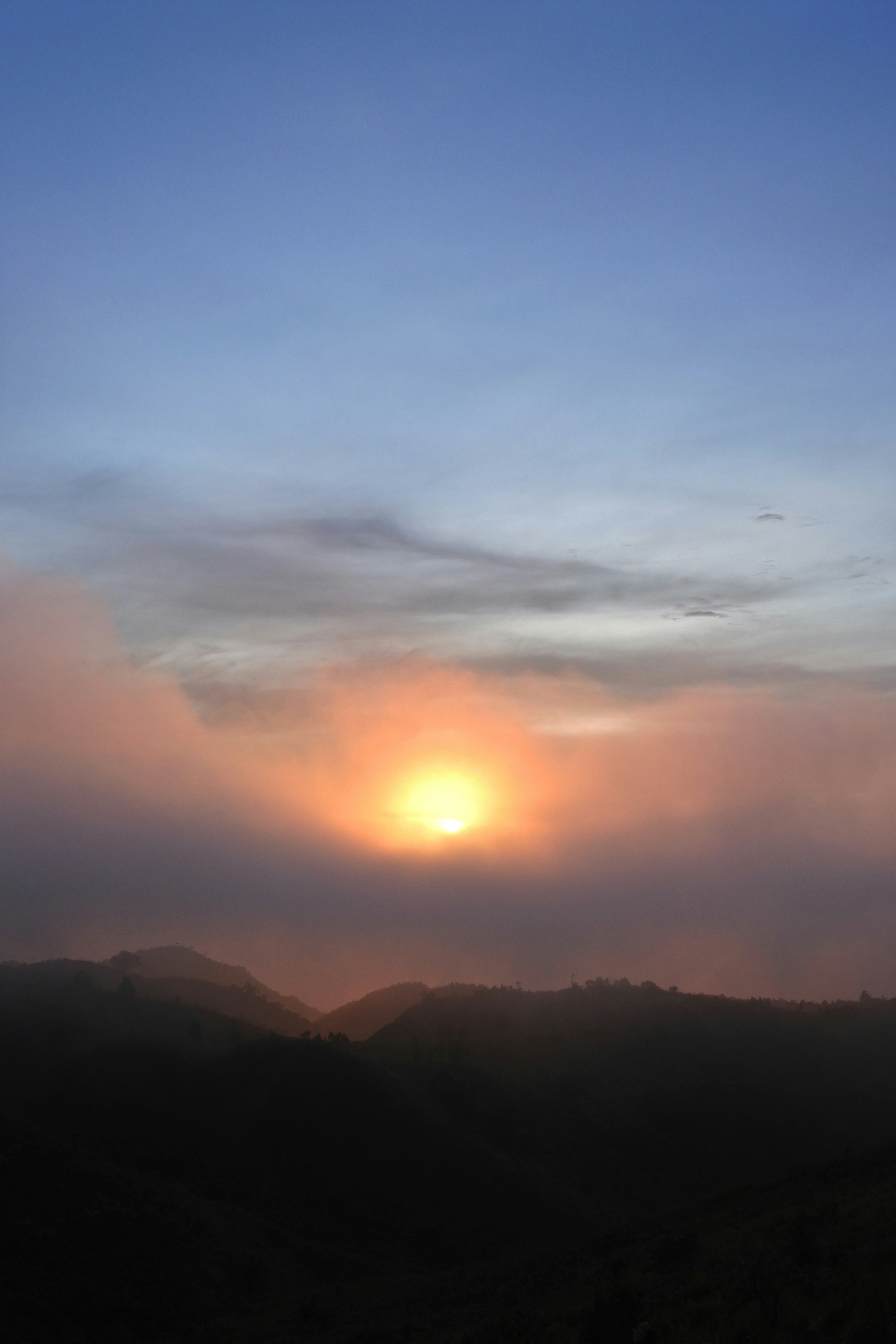 Sunset over rolling hills with a glowing sun breaking through wispy clouds and a hazy horizon.