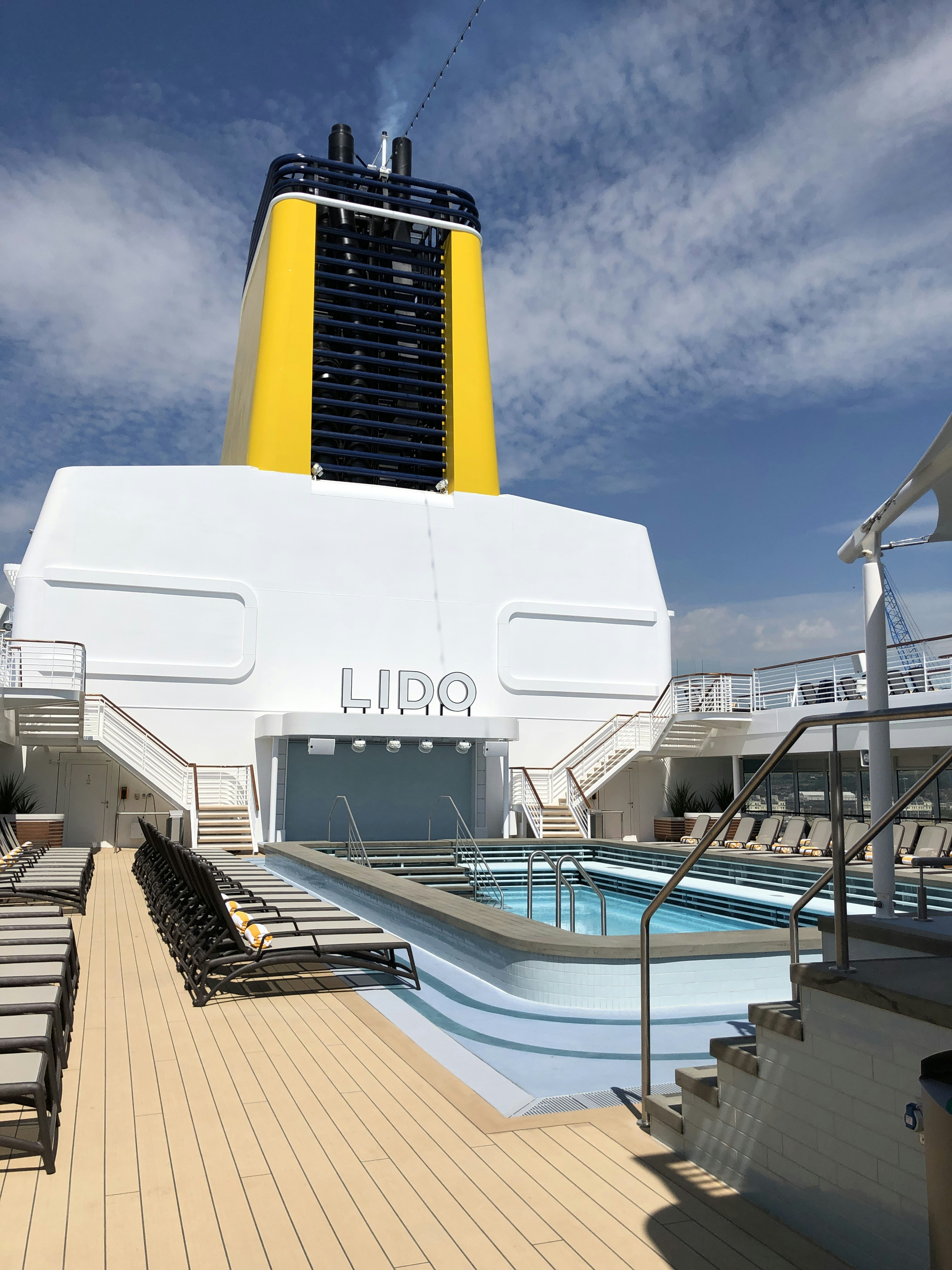Brightly lit Lido deck on a cruise ship featuring a swimming pool and lounge chairs, with a prominent yellow and white structure in the background.