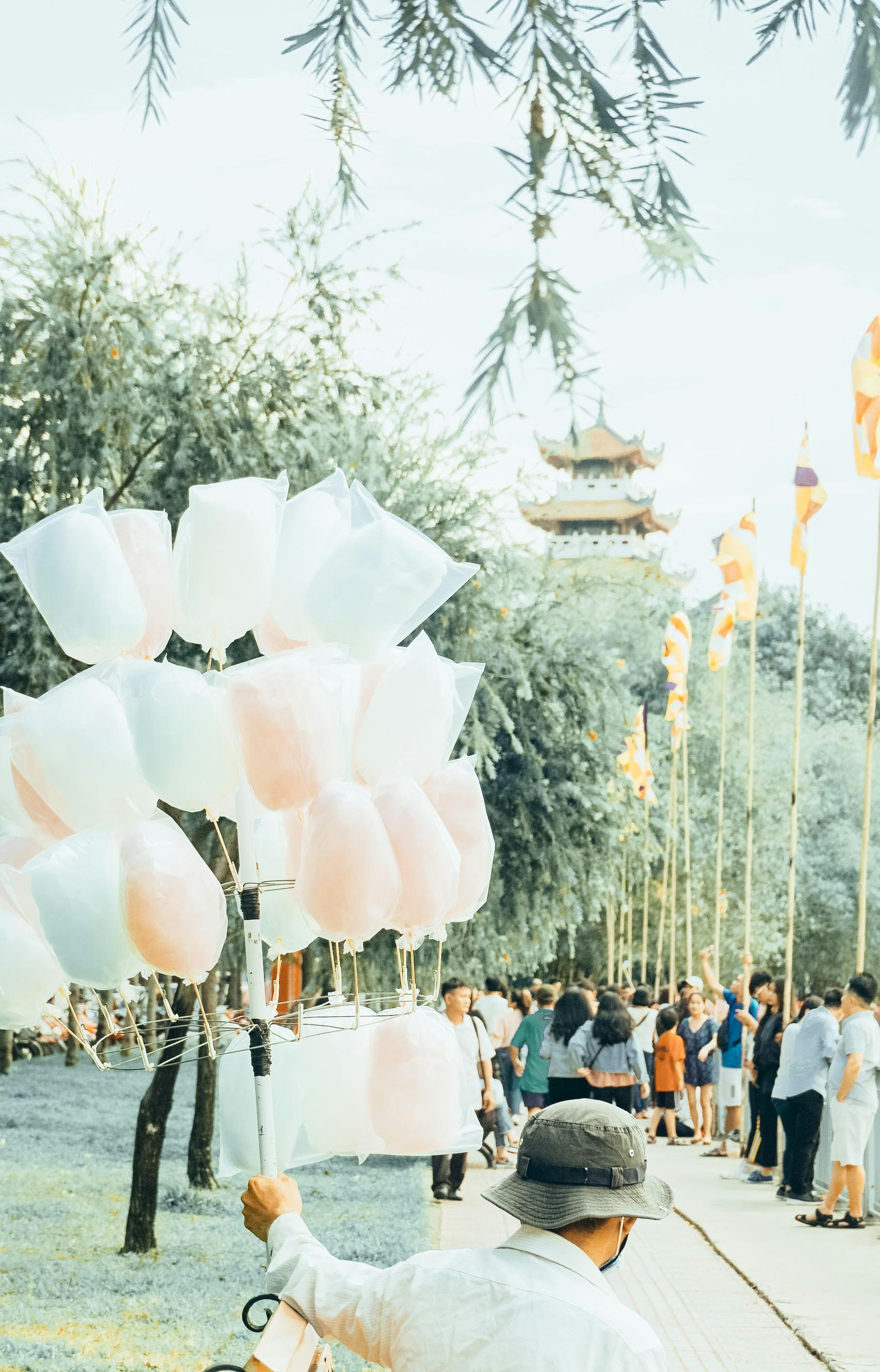 A vendor carries a colorful array of cotton candy while people gather in a lively park setting, framed by trees and flags. The background features a traditional pagoda.