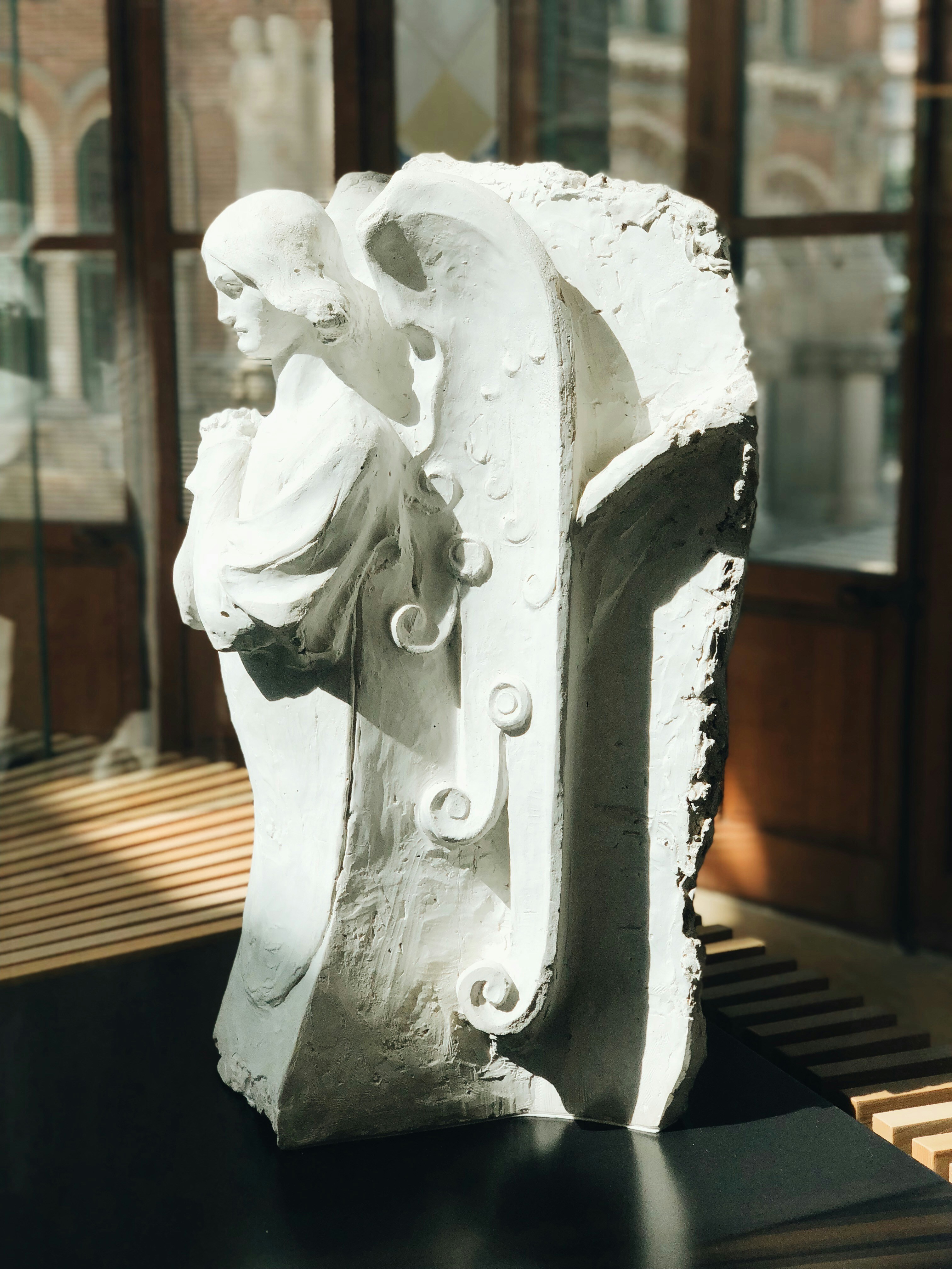 A white sculpture of an angelic figure with flowing robes and intricate details, displayed against a backdrop of wooden architecture.