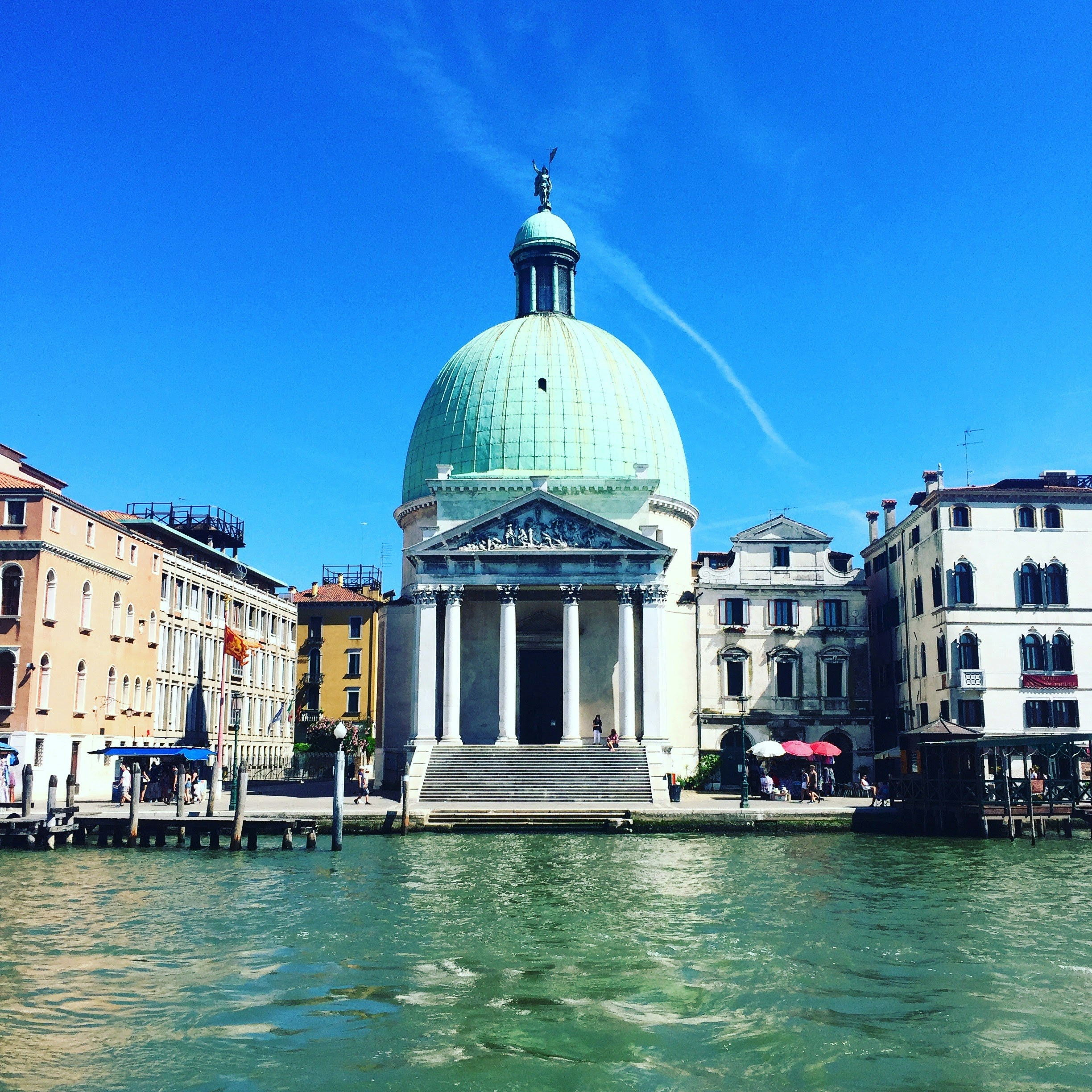 The iconic green dome of Santa Maria della Salute stands majestically against a clear blue sky, framed by historic Venetian architecture along the canal.