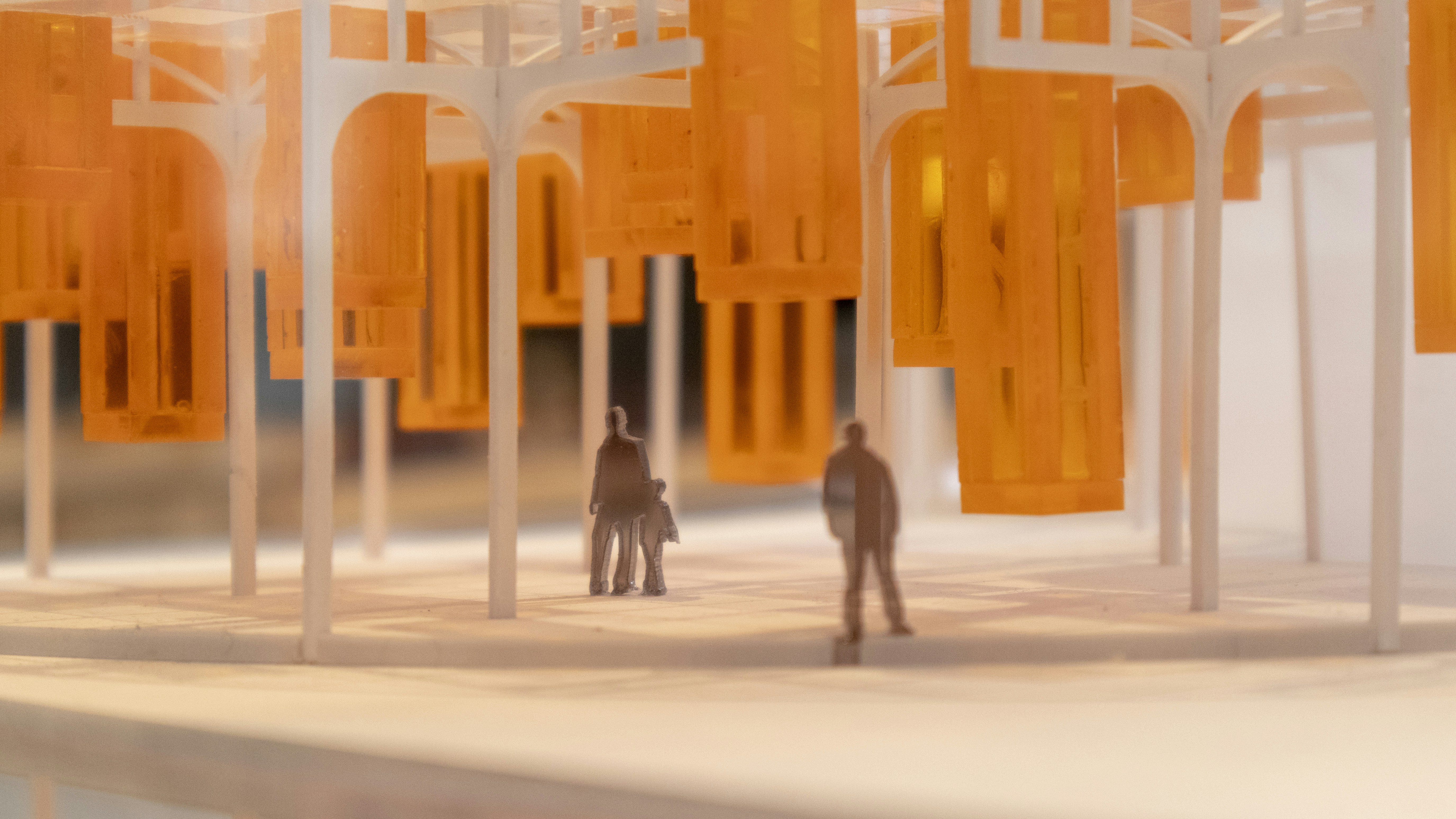 Architectural model featuring orange translucent elements suspended above miniature figures, illustrating spatial dynamics and human interaction.