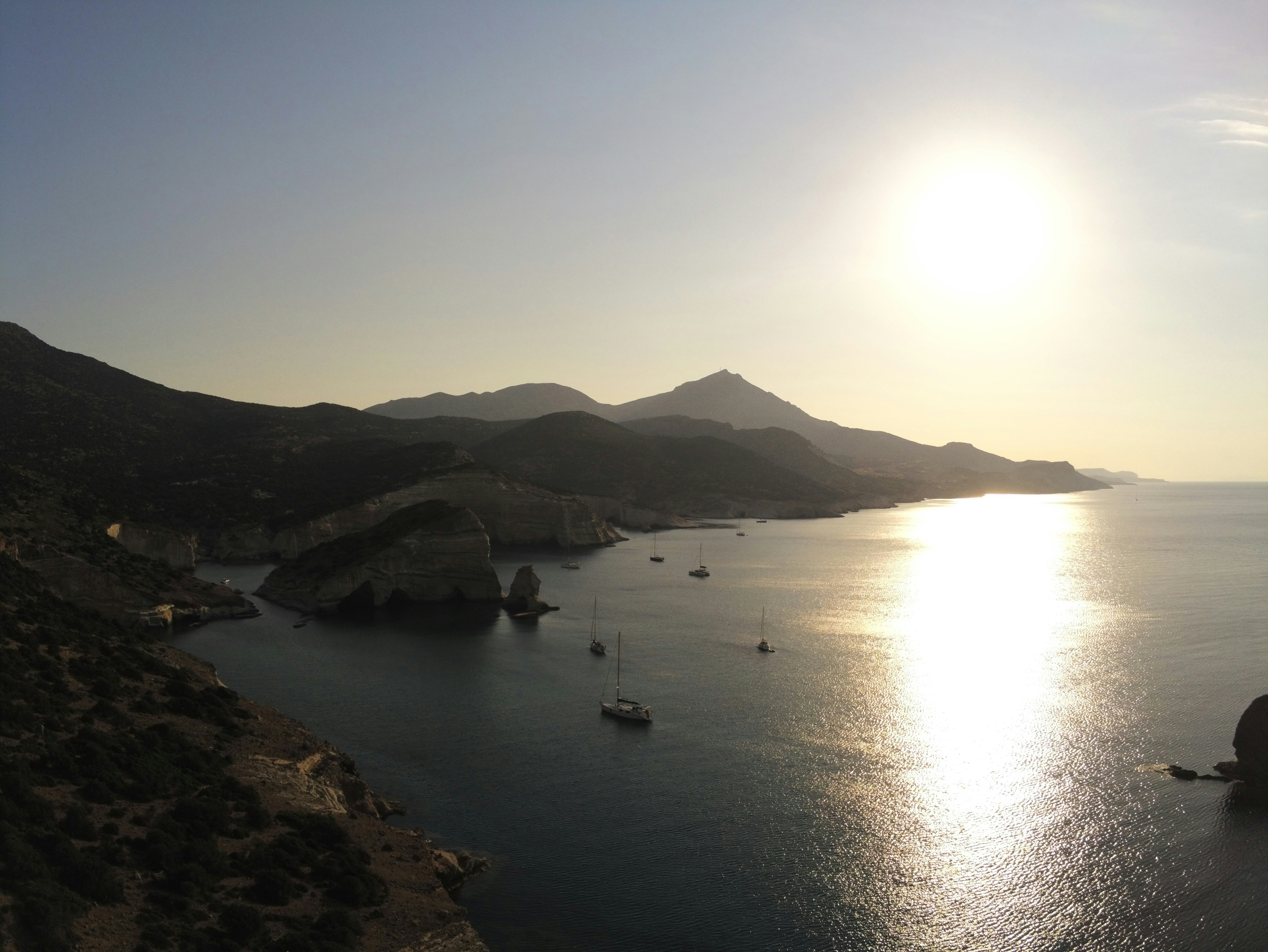 Sunset illuminating calm waters with sailboats anchored near rugged cliffs and distant mountains.