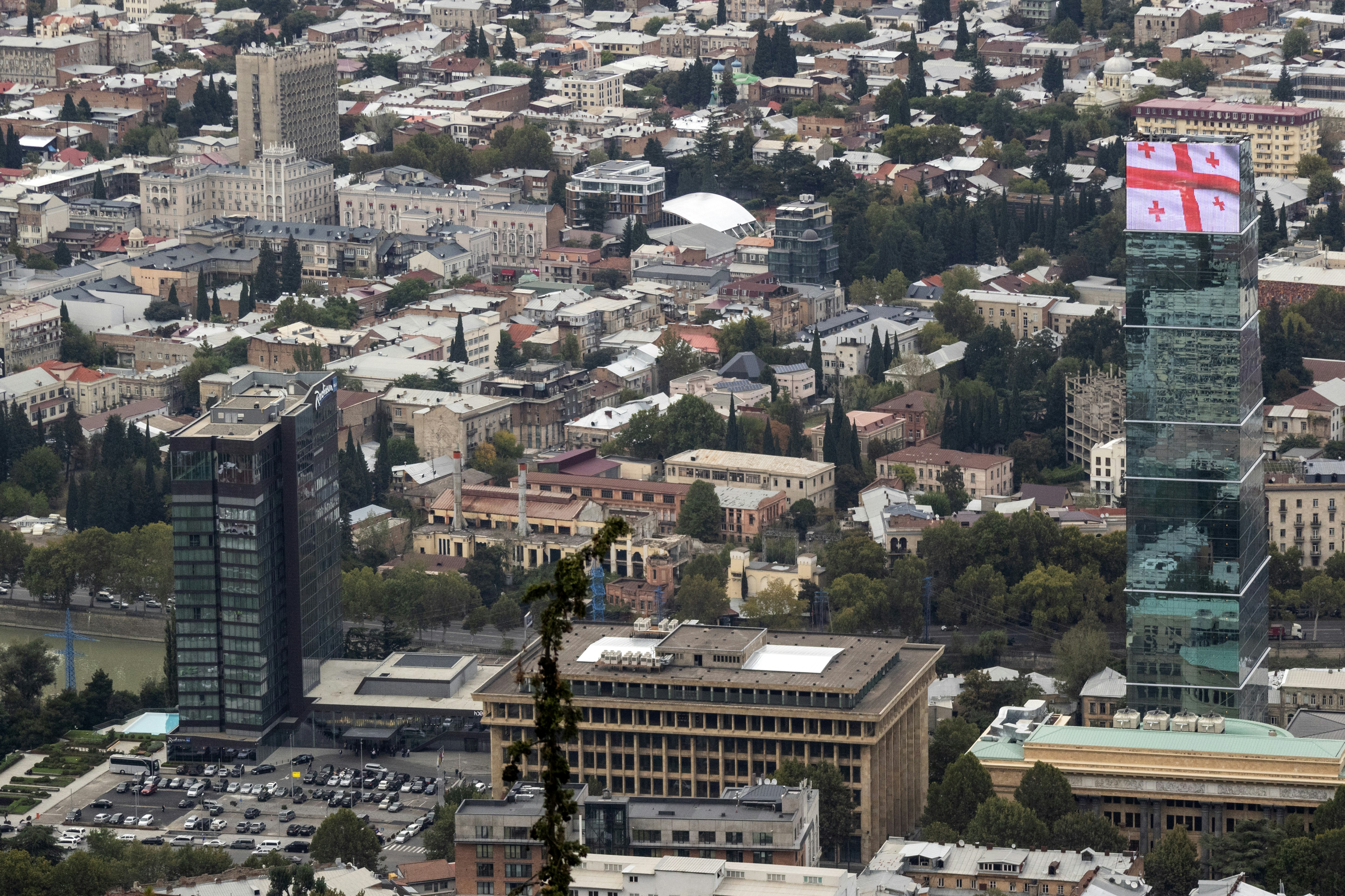 Aerial view of Tbilisi, showcasing a blend of modern skyscrapers and historic buildings amidst lush greenery. The Georgian flag prominently displayed on a high-rise adds a cultural touch.