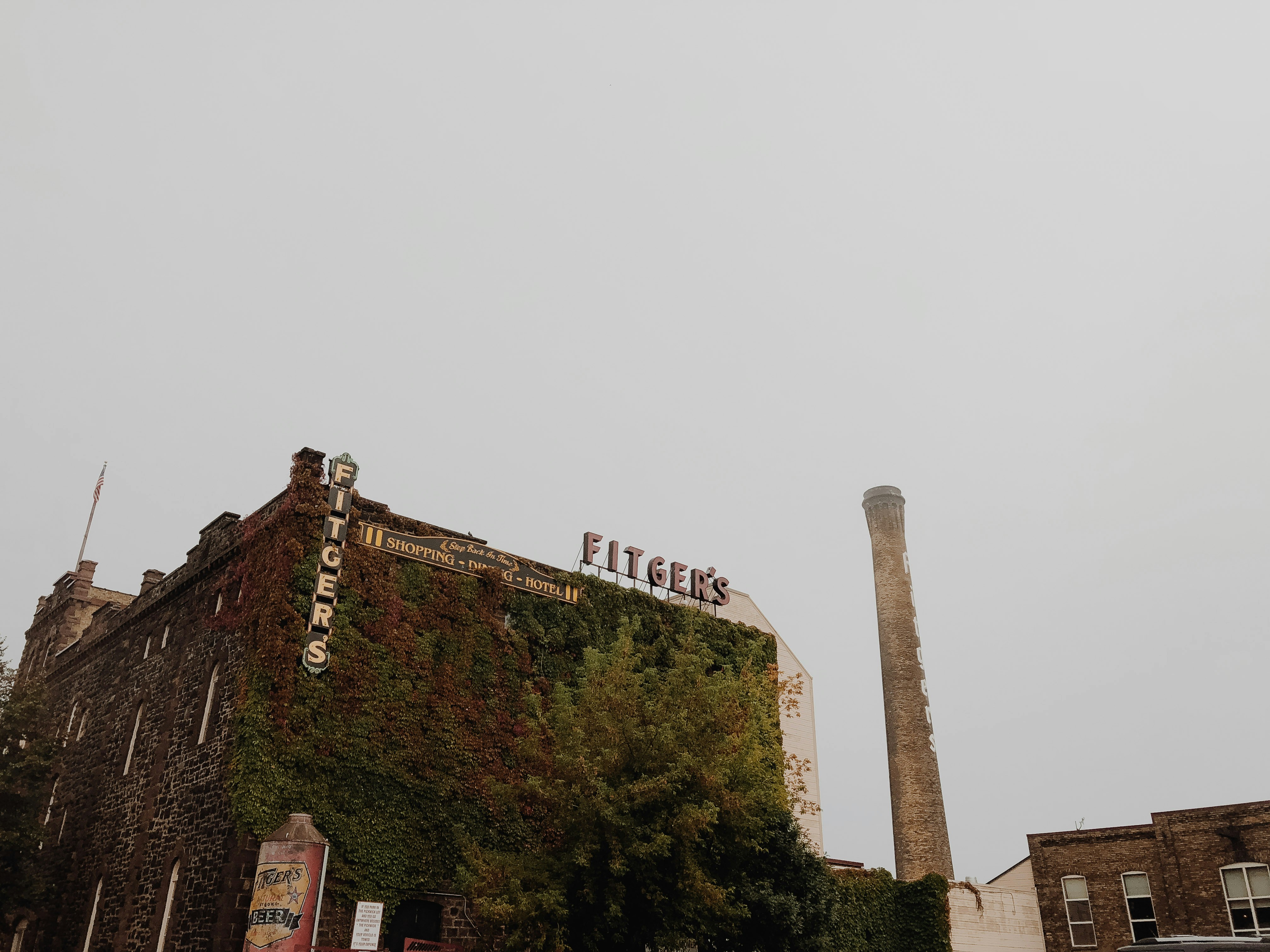 Tall brick building with ivy-covered walls and vintage signage, adjacent to a towering chimney under an overcast sky.