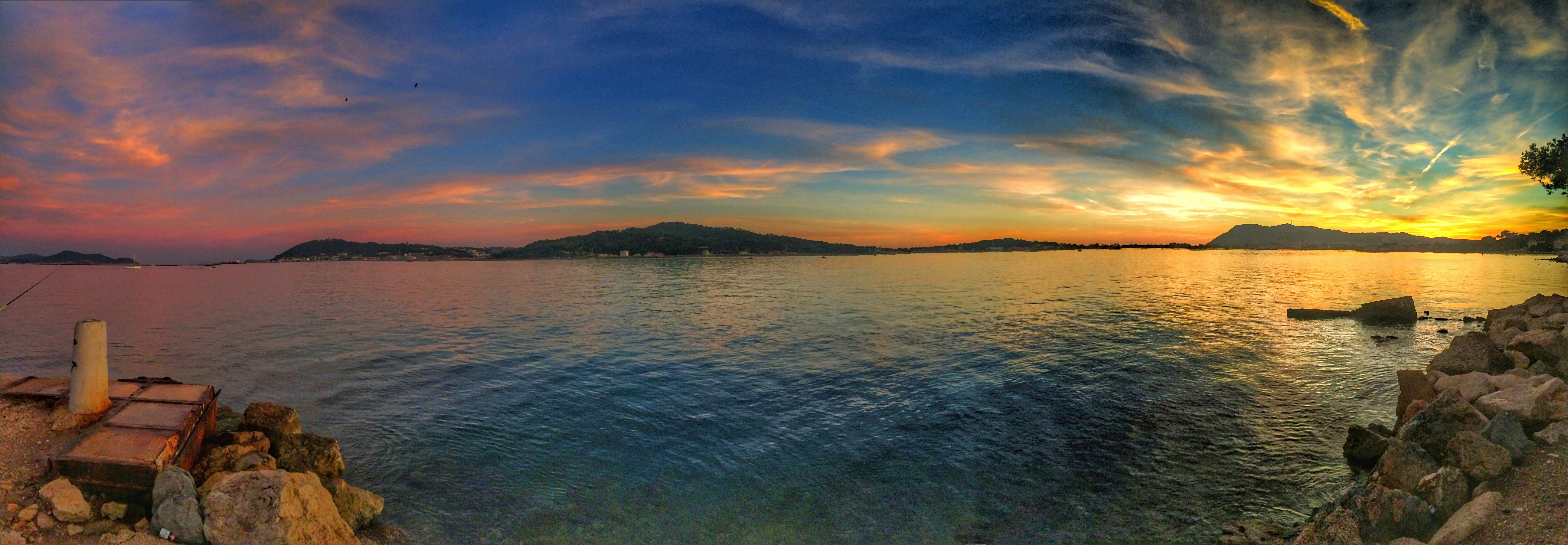 Vibrant sunset over a calm sea with rocky shorelines and silhouetted hills.