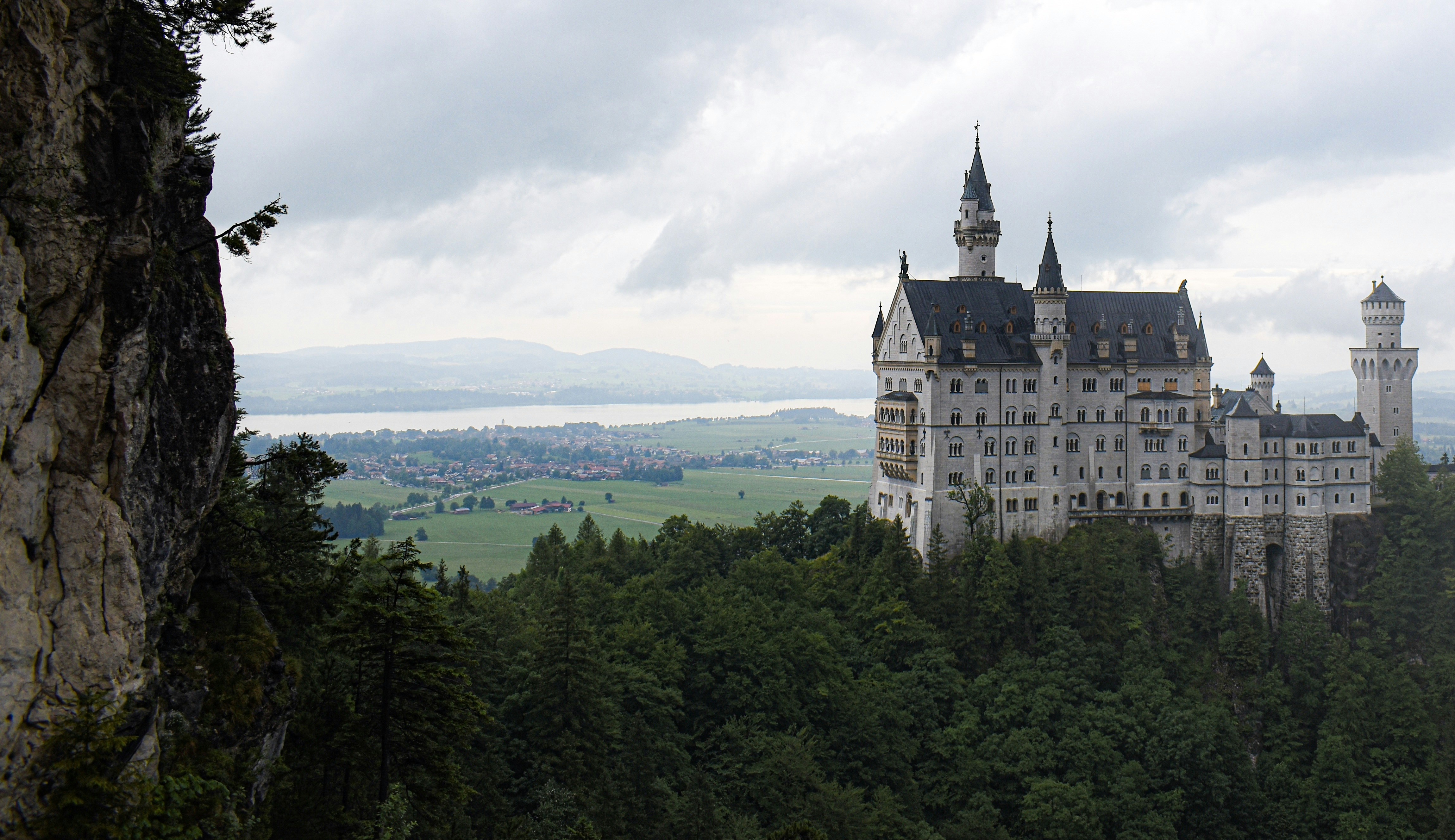 A majestic castle perched on a hillside, surrounded by lush greenery and distant lakes under a moody sky.