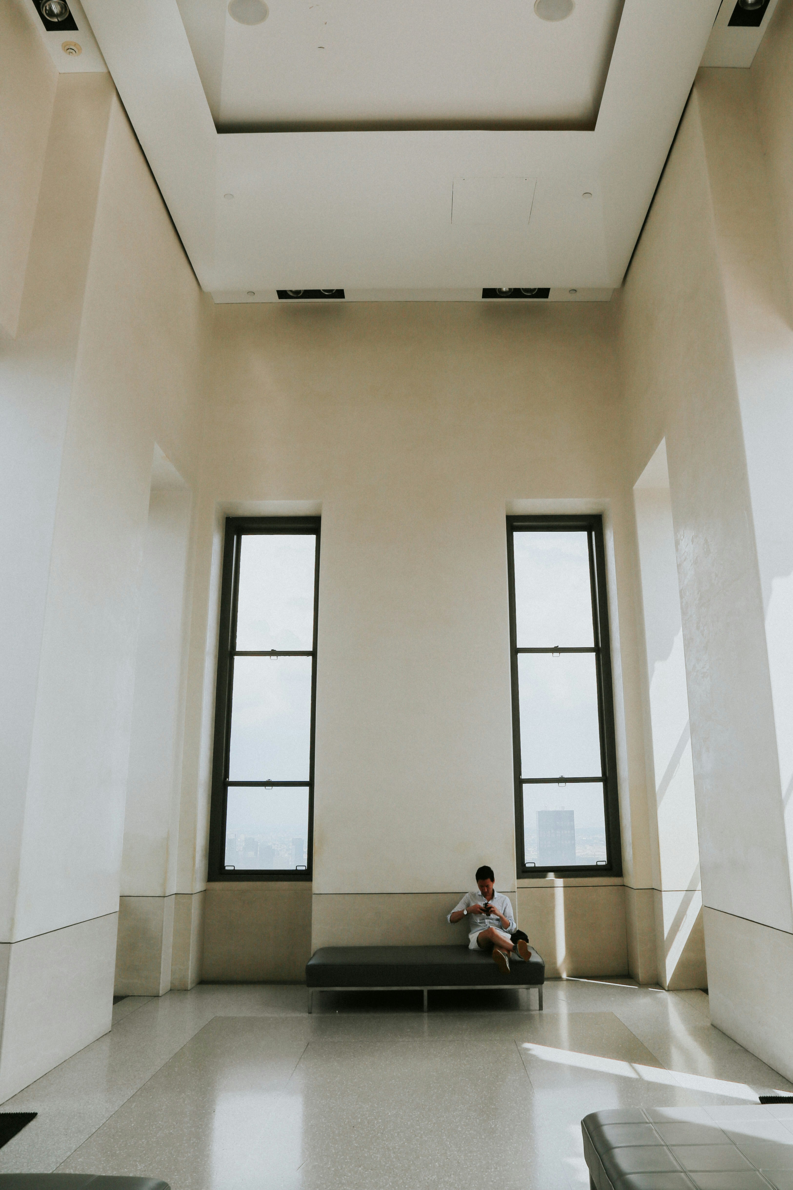 A solitary figure seated on a bench in a spacious, minimalist interior, framed by large windows that let in natural light.