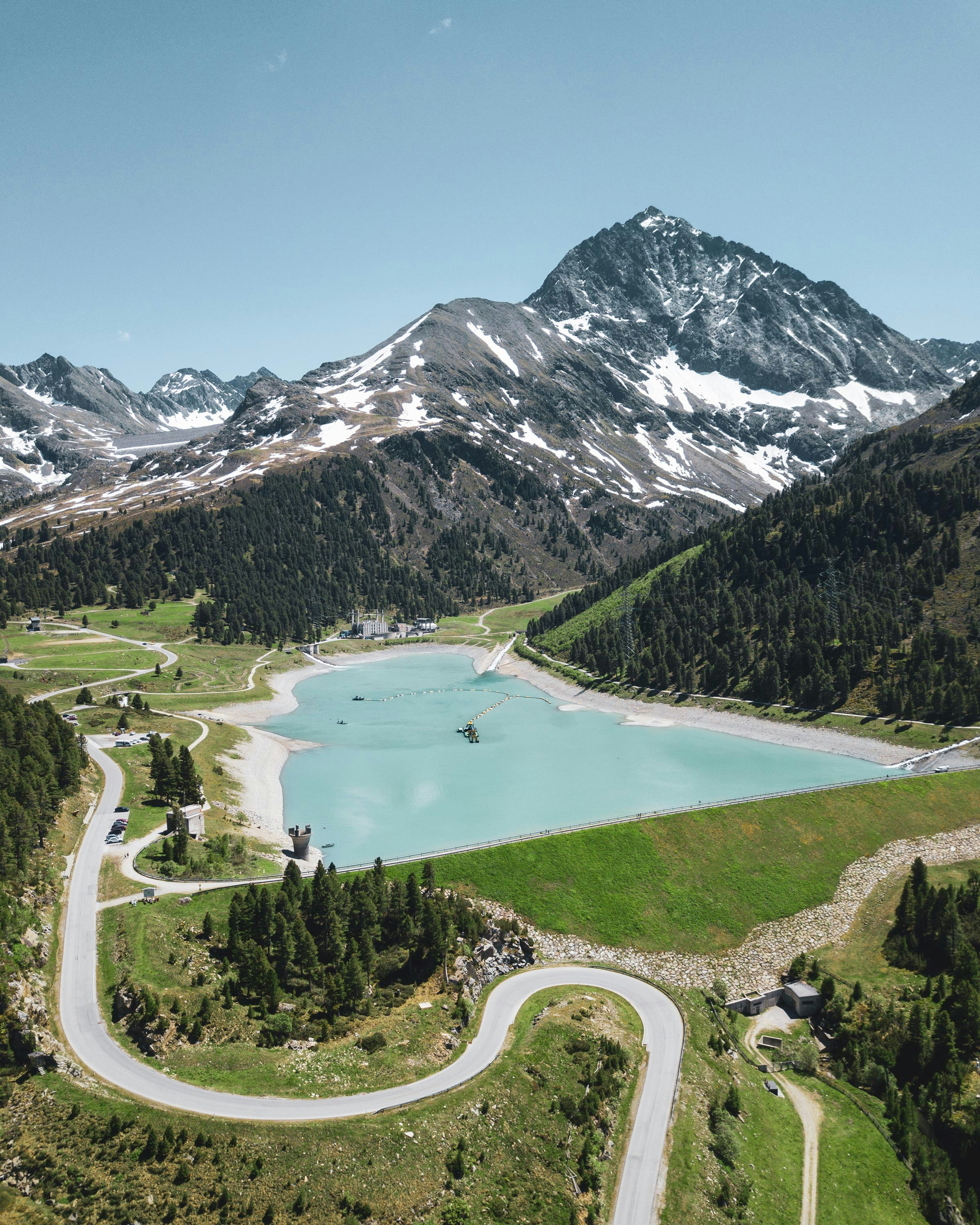 Aerial view of a turquoise reservoir nestled in a mountainous landscape with winding roads and snow-capped peaks.