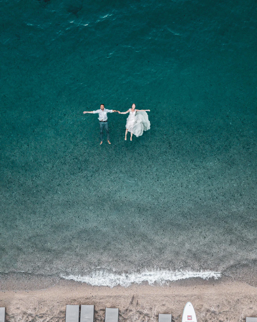A bride and groom float hand in hand in clear teal water near a sandy beach. The shoreline, loungers, and a small surfboard appear at the bottom edge.
