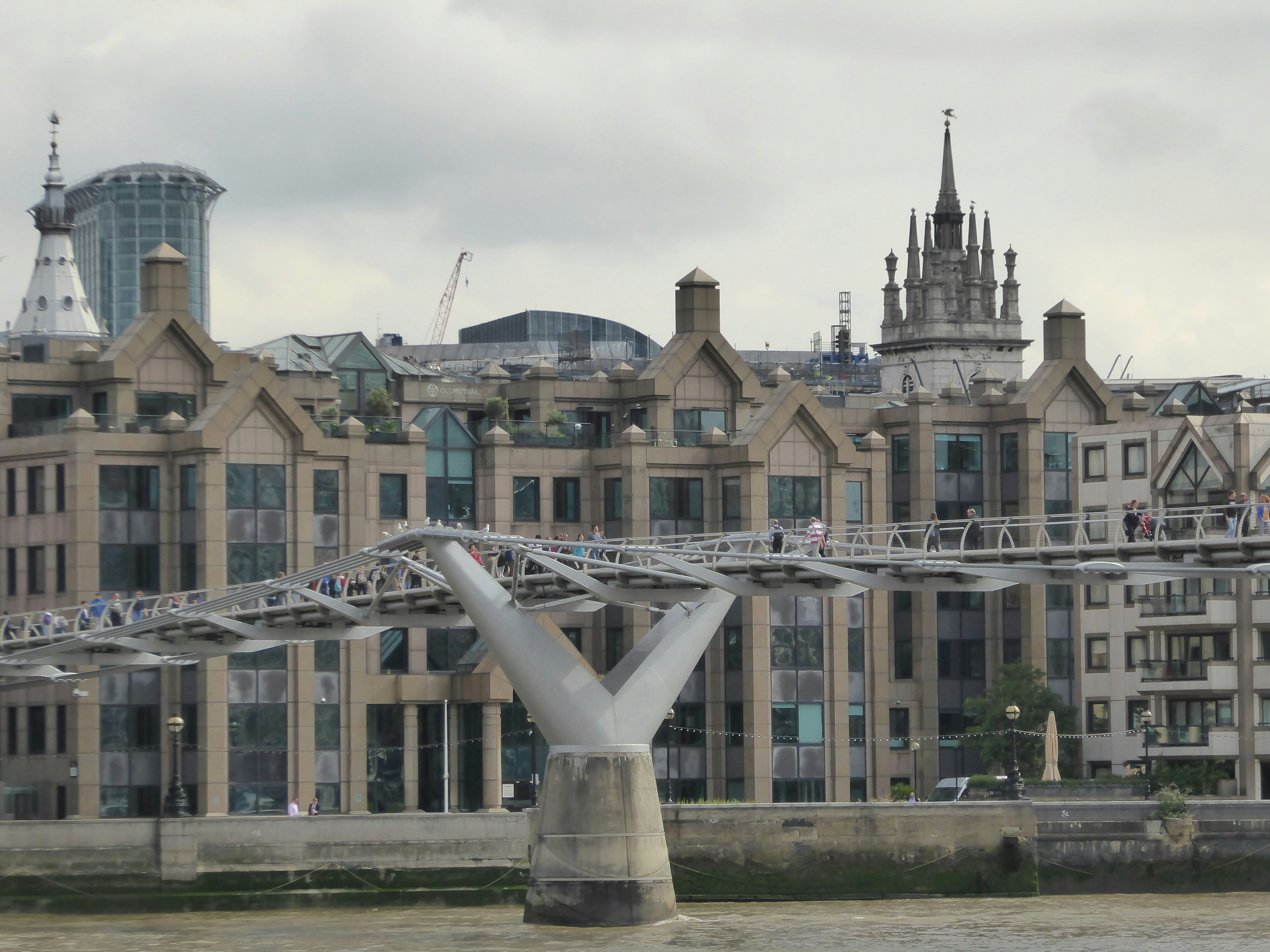 Contemporary pedestrian bridge spans over a river with classic architecture in the background under a cloudy sky.