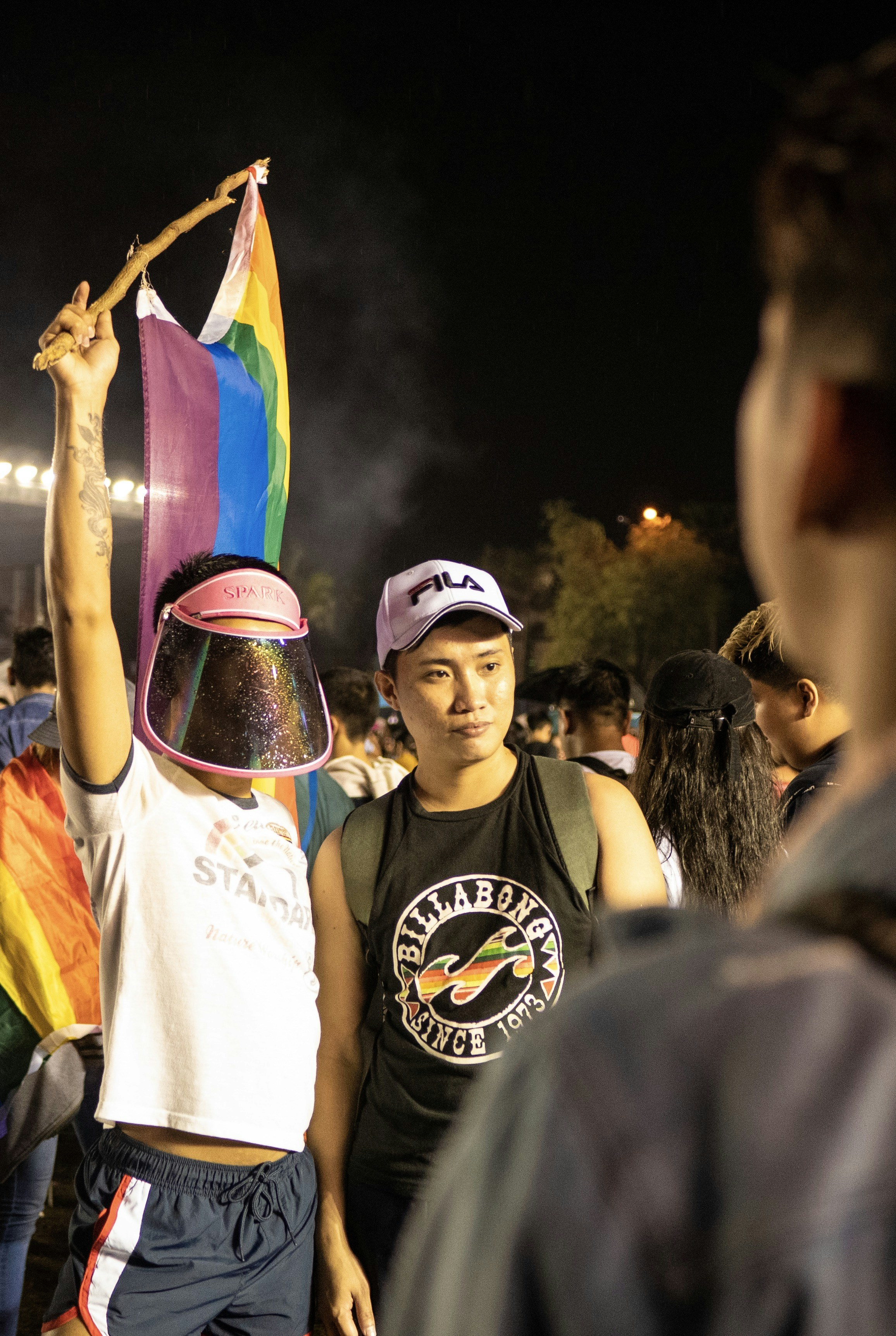 Two individuals at a lively event, one holding a rainbow flag and wearing a visor, while the other sports a casual outfit. The atmosphere exudes celebration and unity.