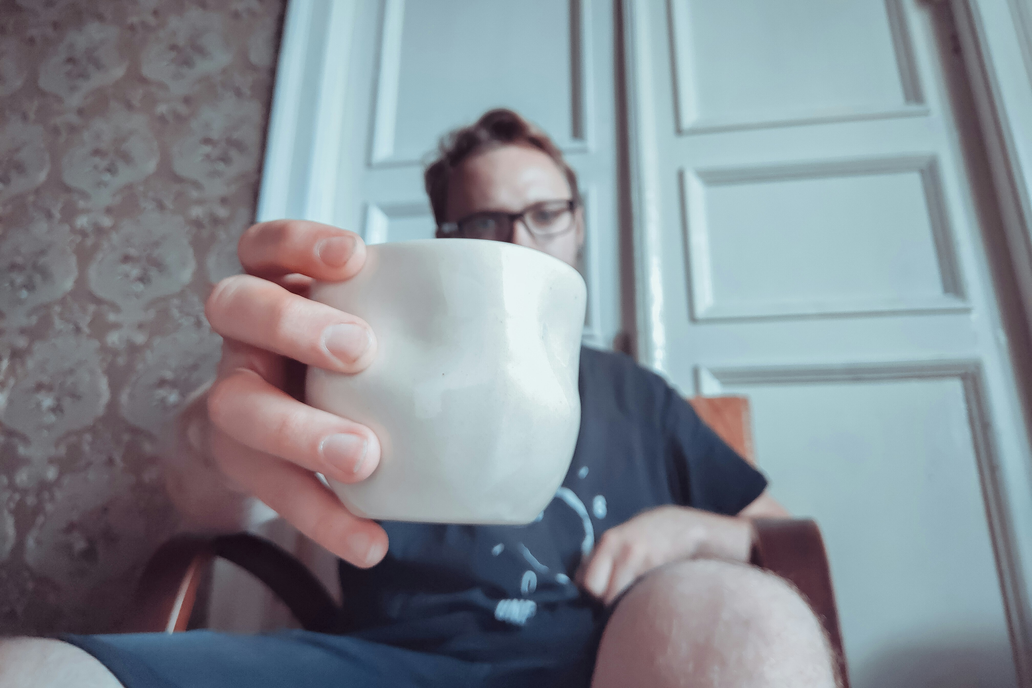 A close-up photograph of a hand holding a white cup toward the camera, with a blurred figure in a dim, domestic setting.