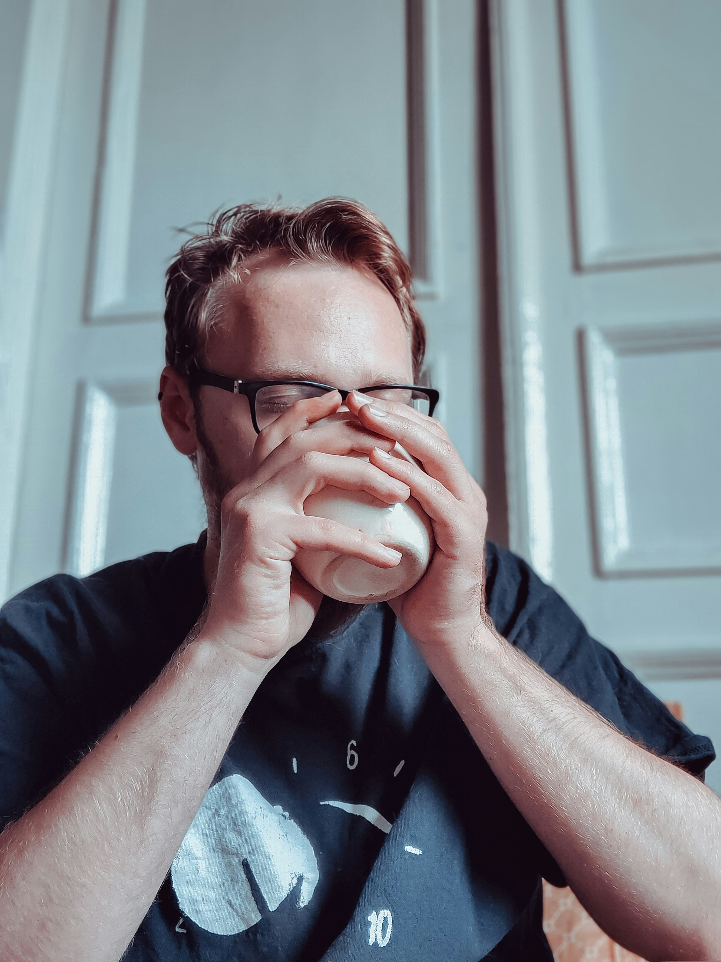 Portrait photo of a man with glasses drinking from a white cup, set against pale paneled doors in cool indoor light.