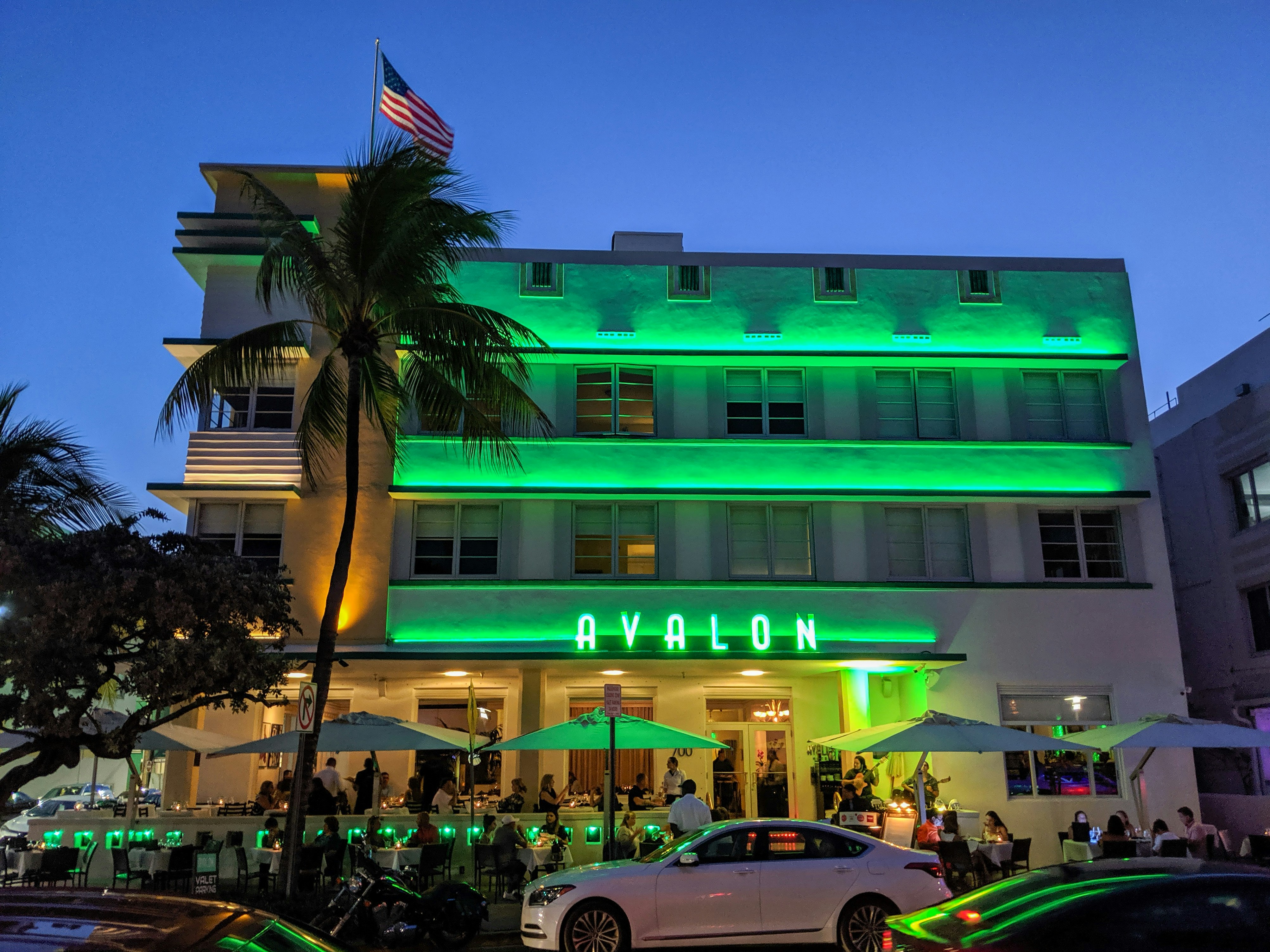 Art Deco hotel illuminated in vibrant green with a lively outdoor dining scene under umbrellas. American flag waves above.