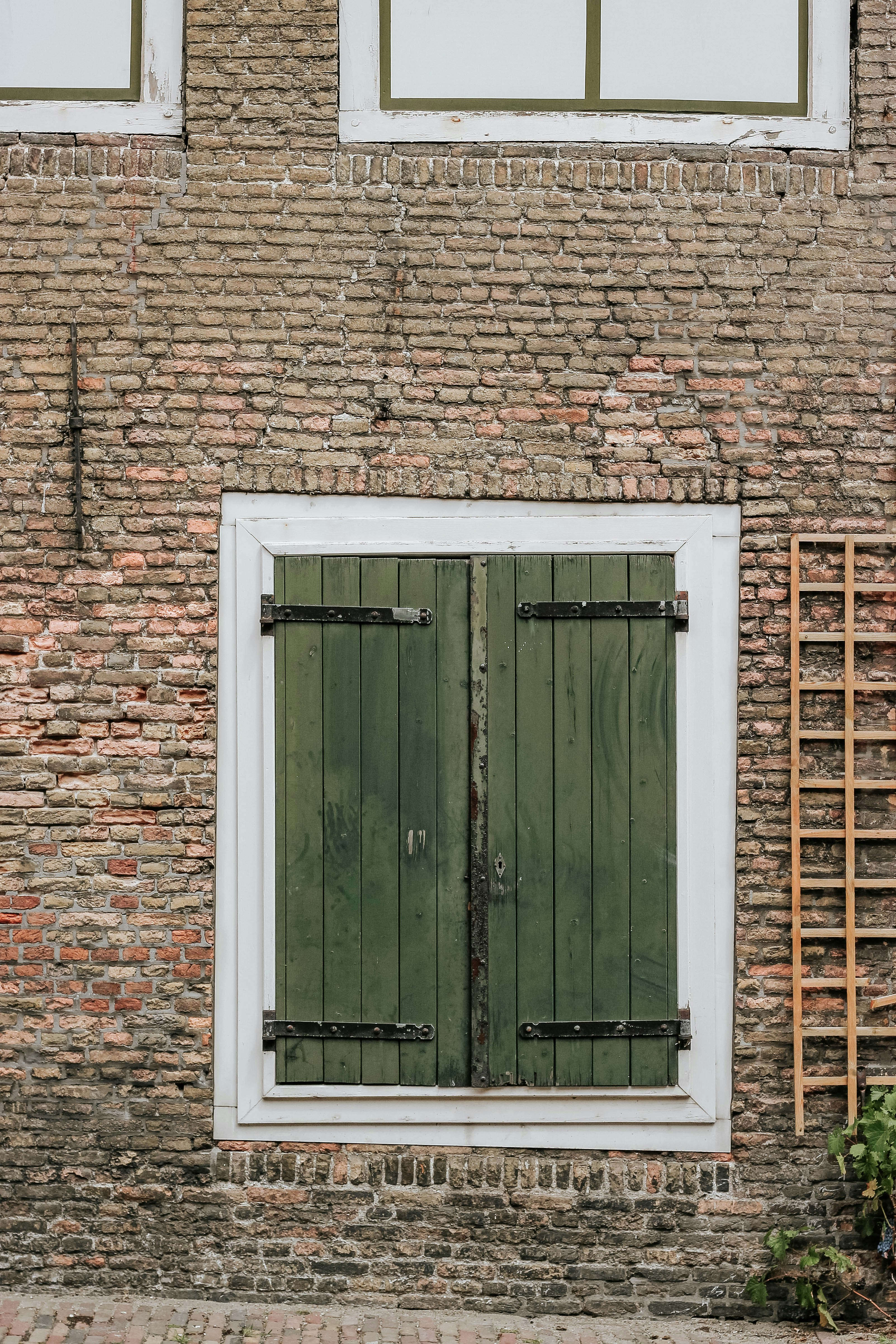 Weathered green shutters on a brick wall, showcasing the texture and history of the building. A wooden trellis adds a touch of nature.