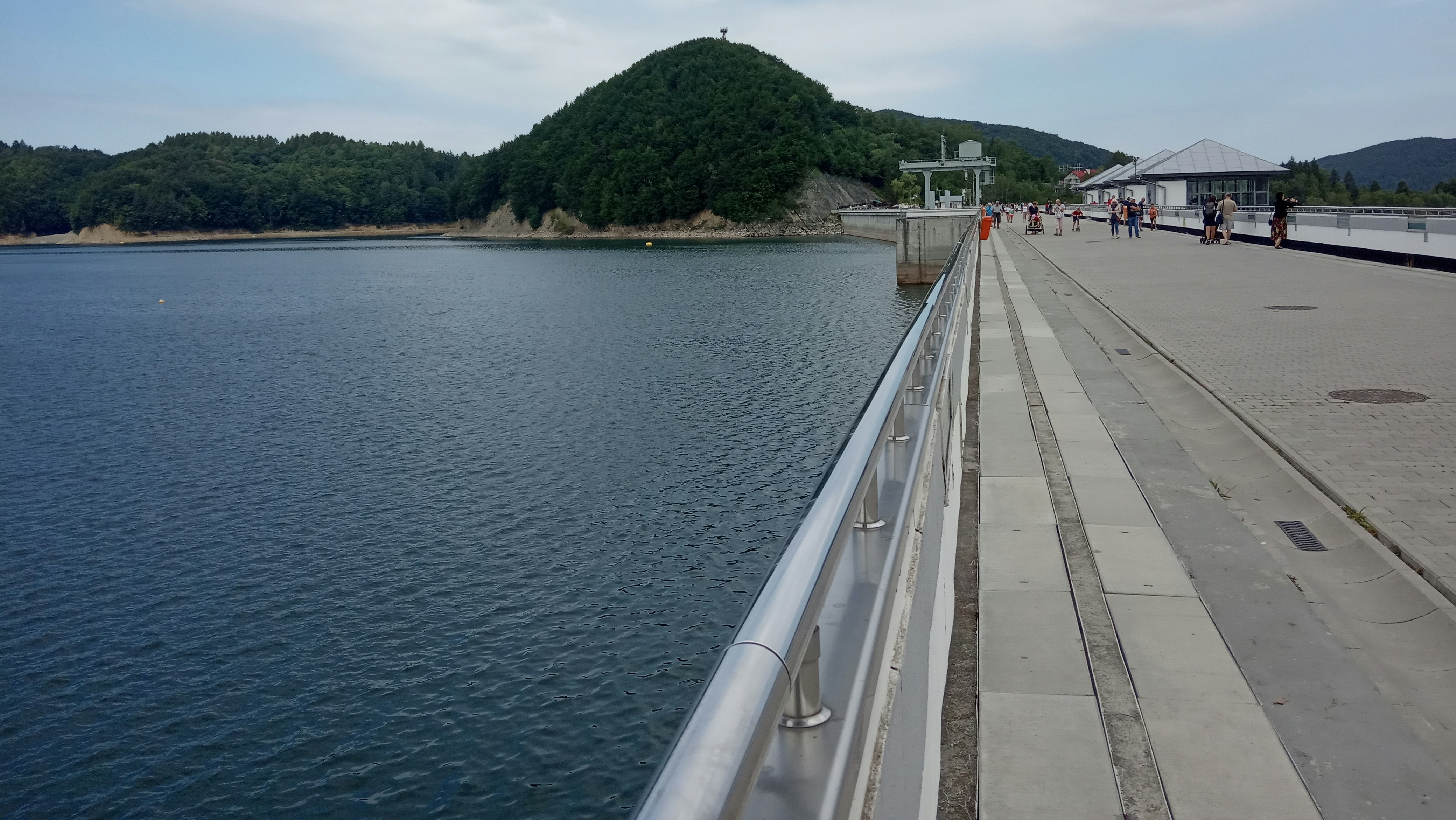 Wide waterfront promenade with a metal railing stretching toward distant hills. Calm blue water sits to the left as pedestrians walk along the concrete path.