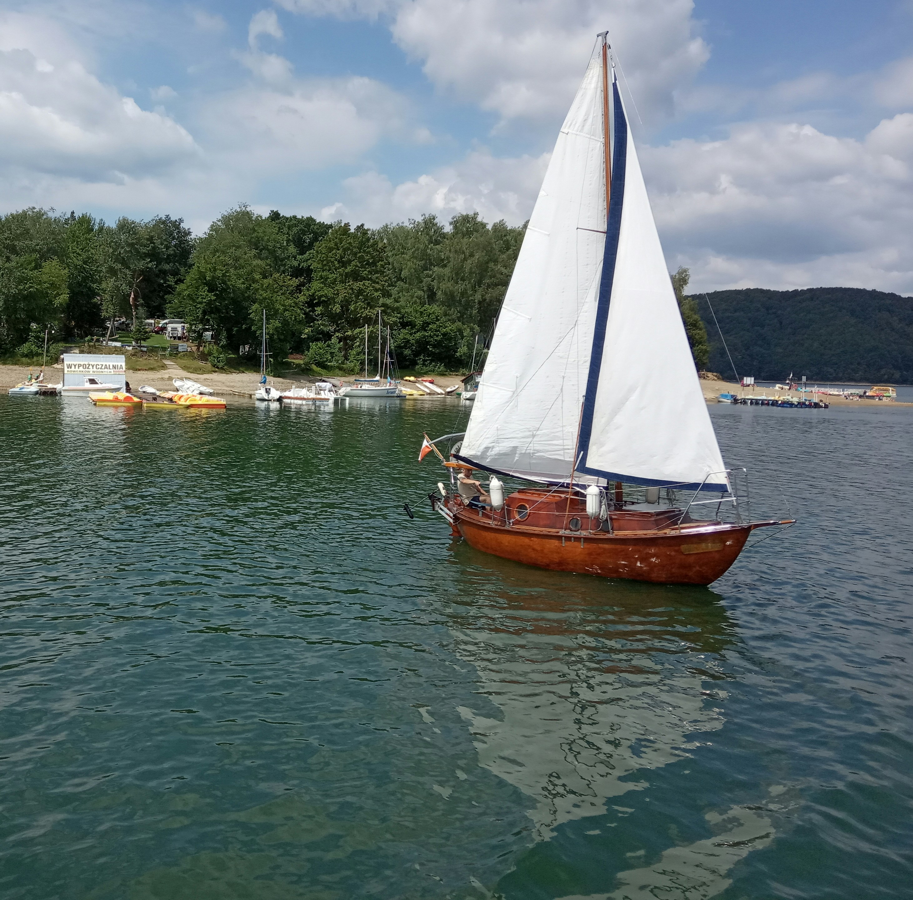 Photograph of a wooden sailboat with white sails gliding on a calm lake, bordered by a tree-lined shore and distant hills.