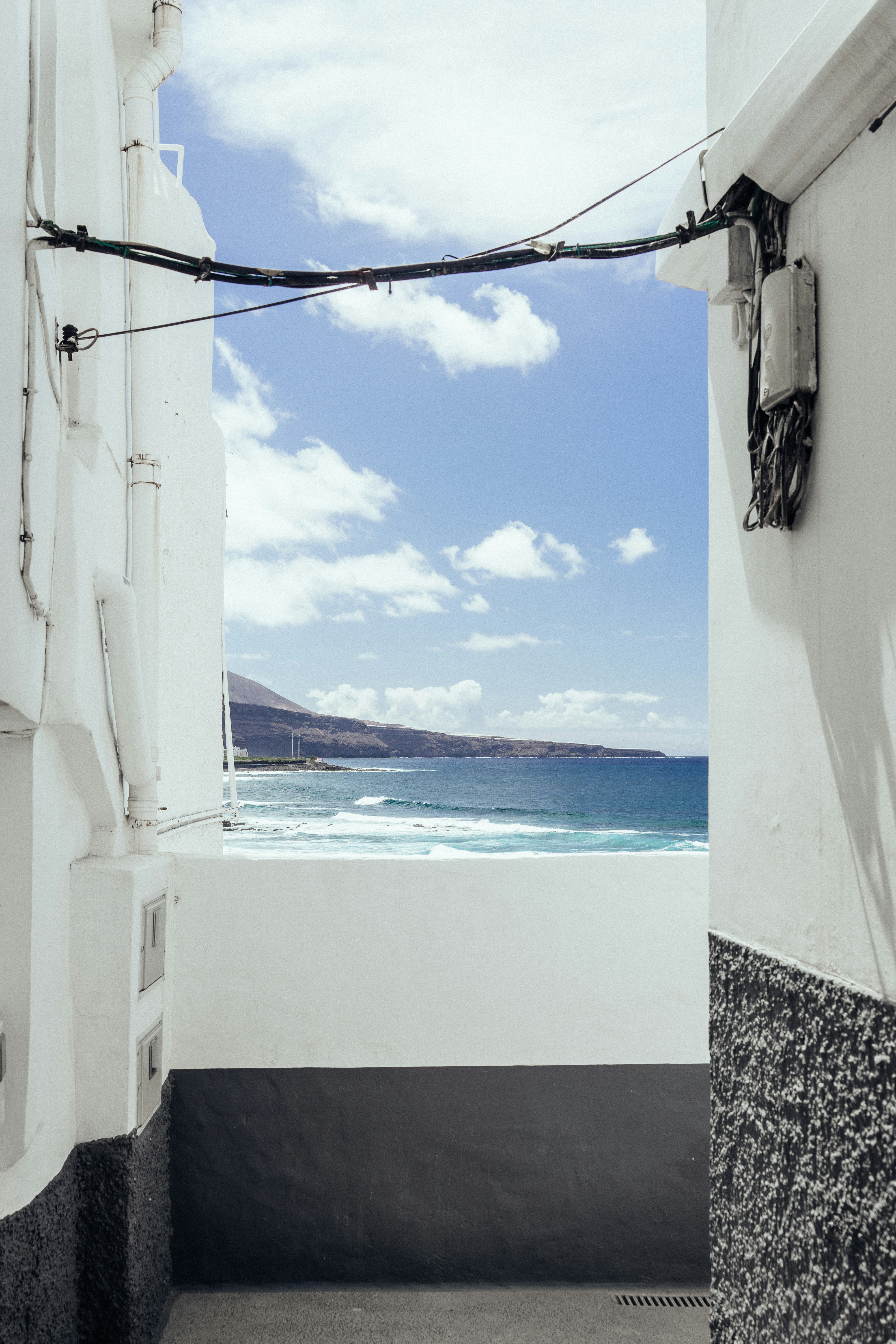 A narrow passageway reveals a tranquil coastal scene with gentle waves and distant mountains, framed by whitewashed walls and contrasting textures.