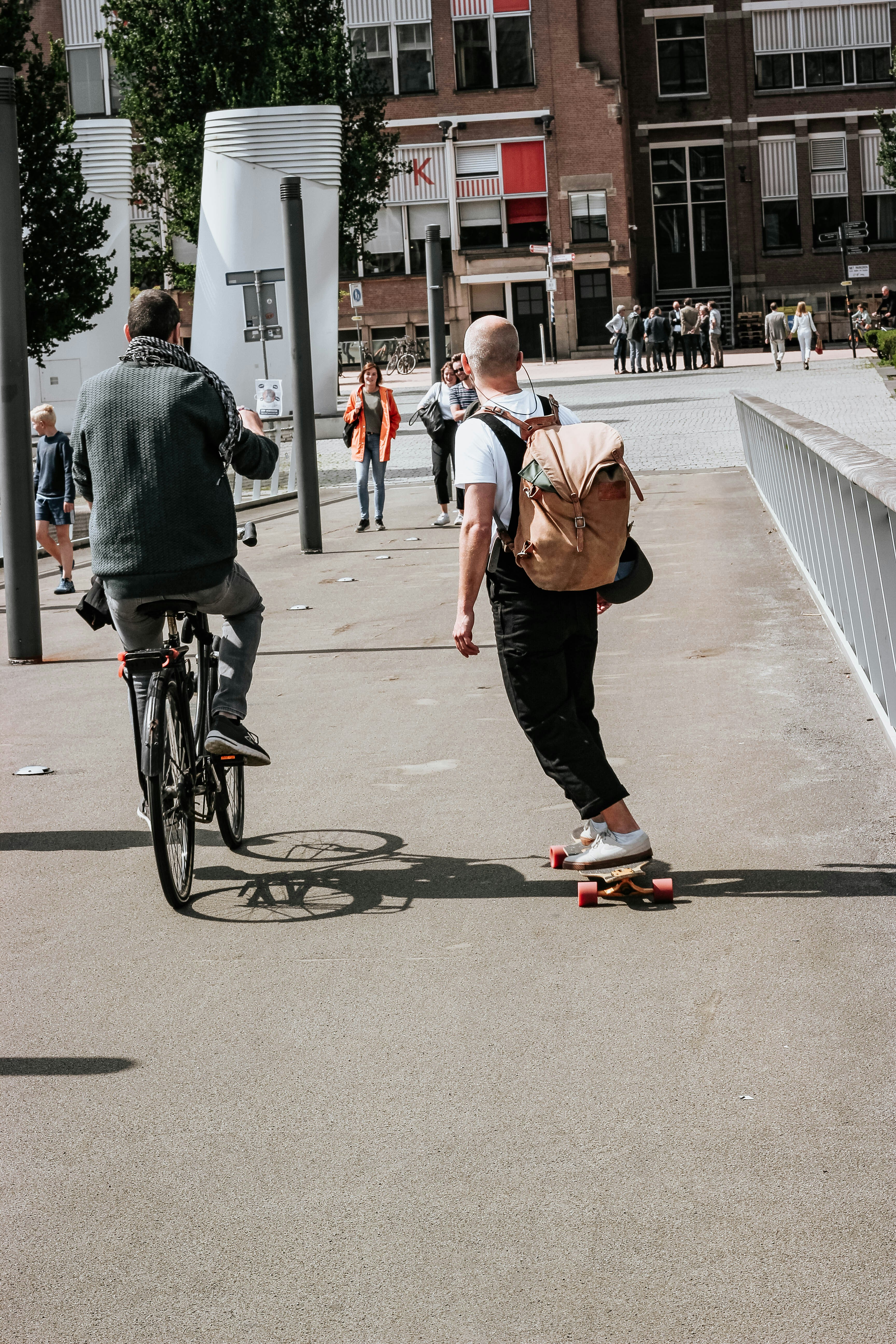 A cyclist and a skateboarder navigate a bustling city pathway, showcasing urban mobility and leisure. The scene captures a vibrant mix of movement and city life.