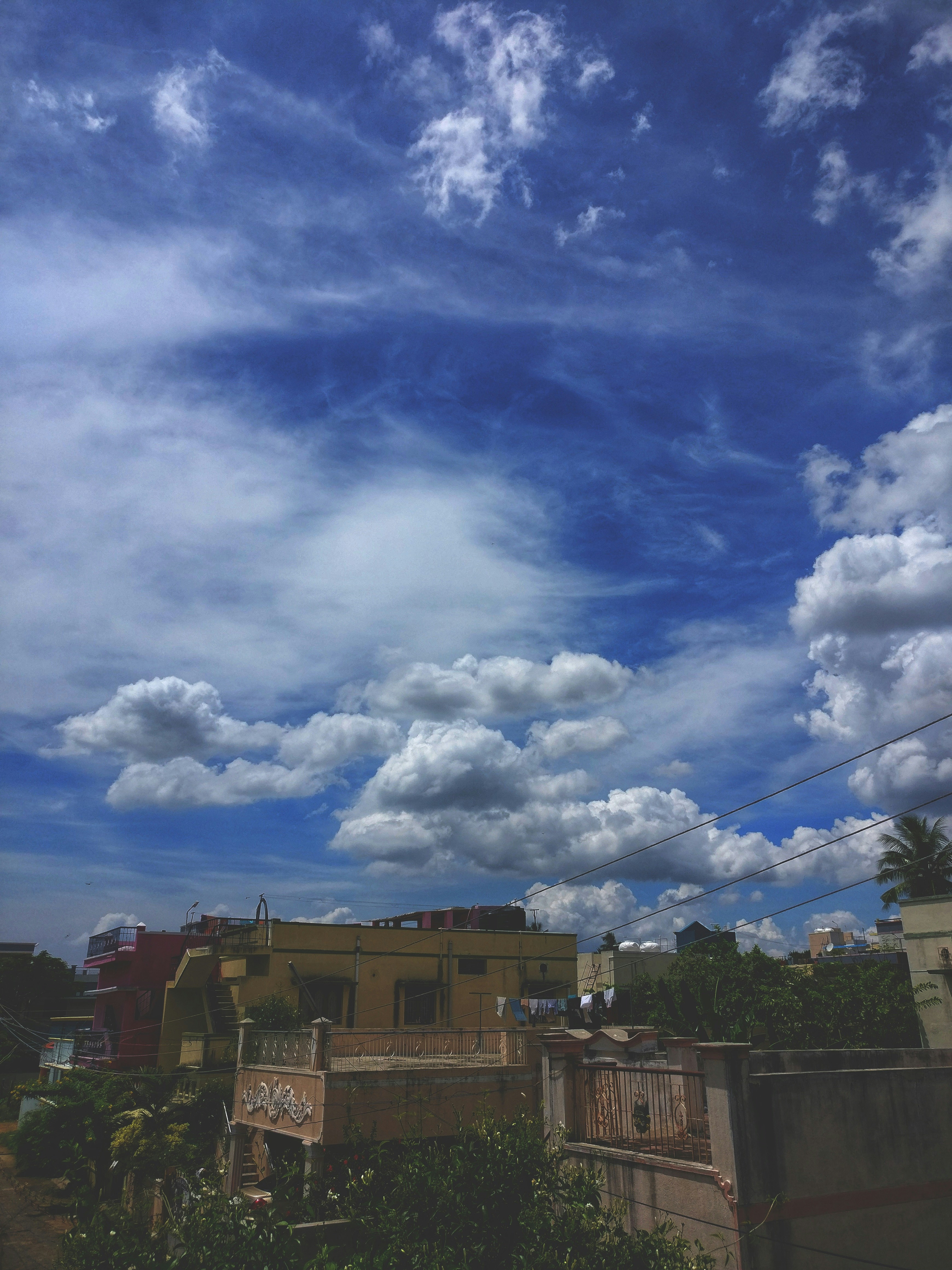 Fluffy clouds drift across a vibrant blue sky above colorful buildings in an urban setting. The scene captures a tranquil moment amid city life.