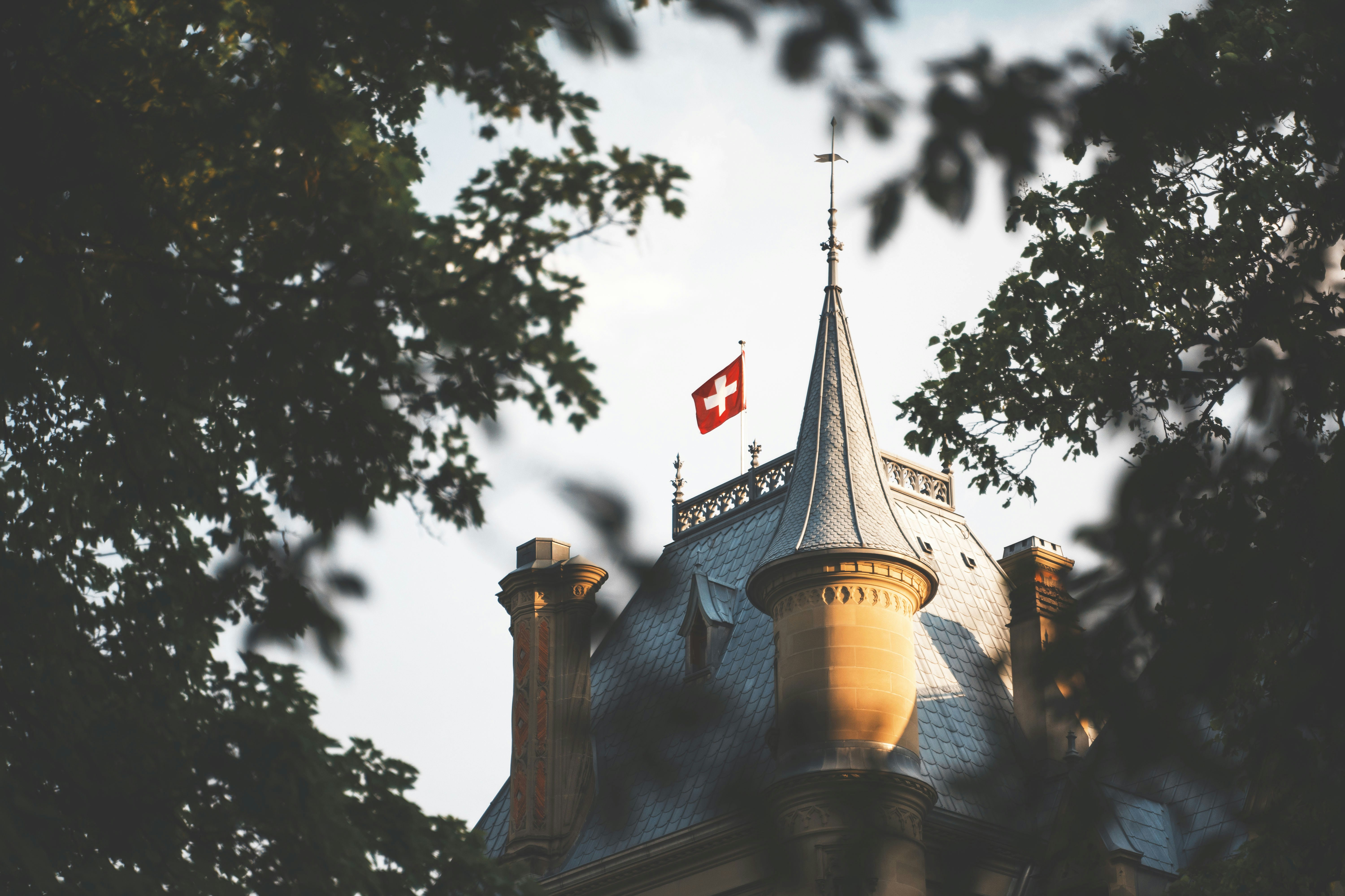 Historic tower with a Swiss flag framed by lush green foliage under a soft evening light.