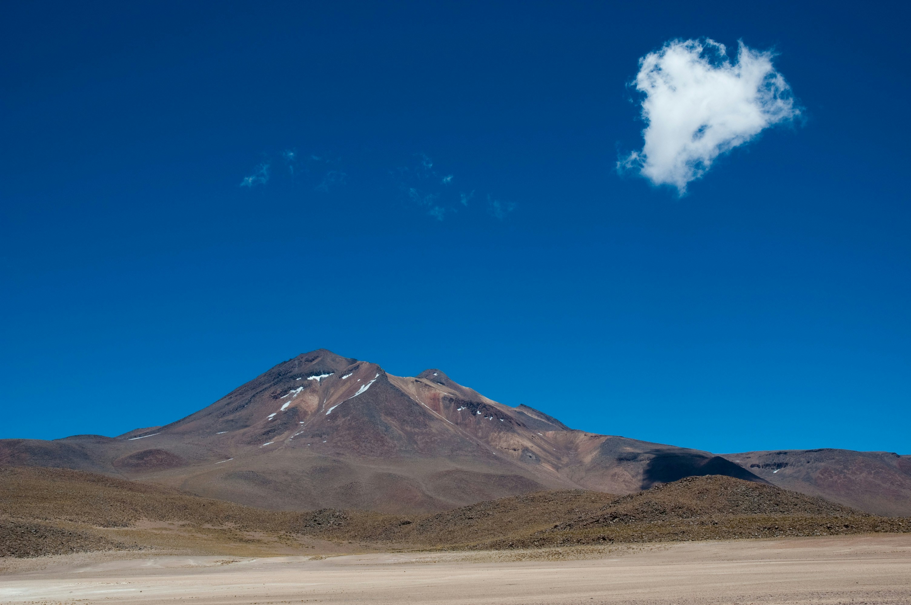 Vast arid landscape featuring a prominent mountain under a clear blue sky, with a solitary cloud drifting above.