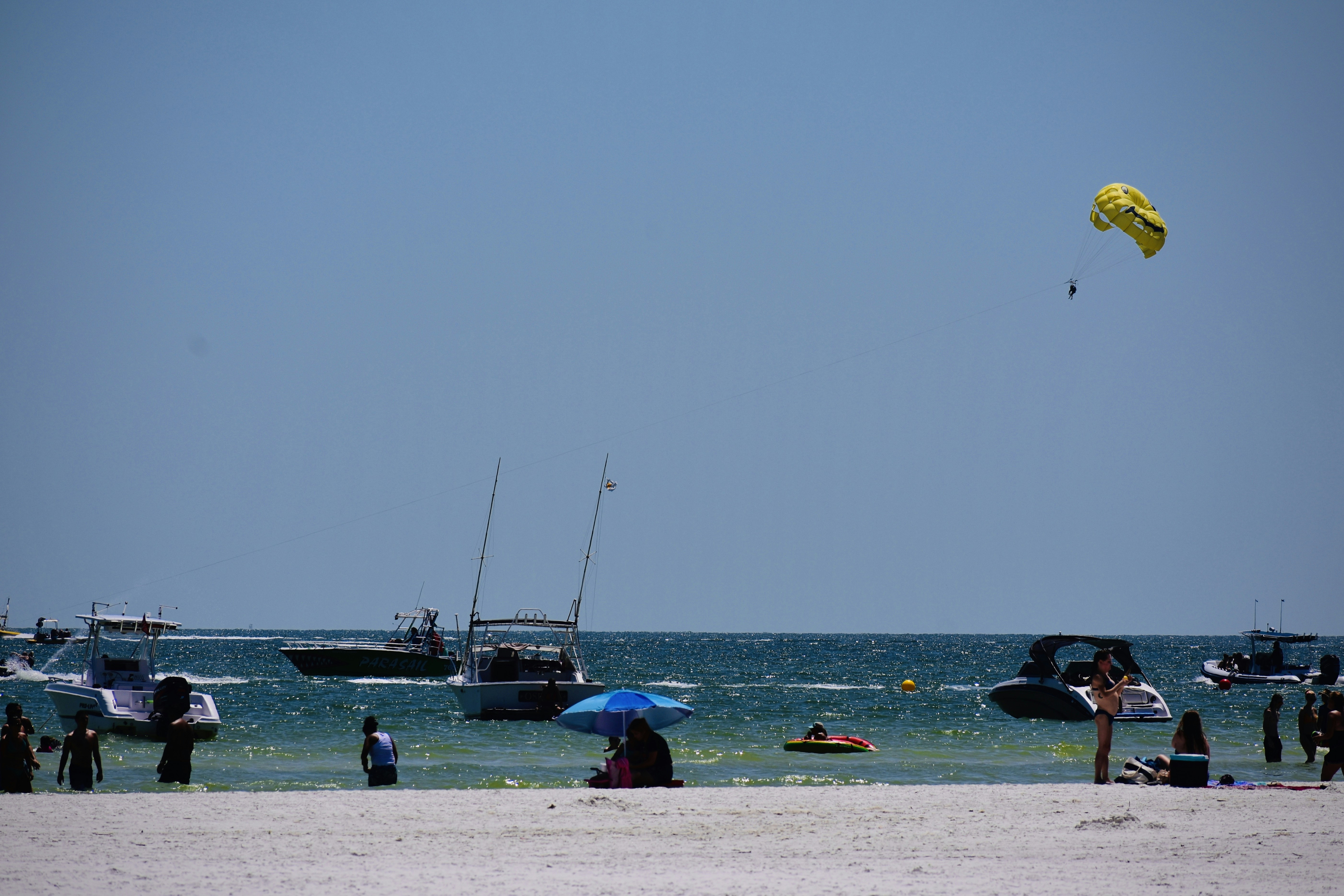 Beachgoers enjoying a sunny day as boats navigate the shimmering waters, with a parachute gliding overhead.
