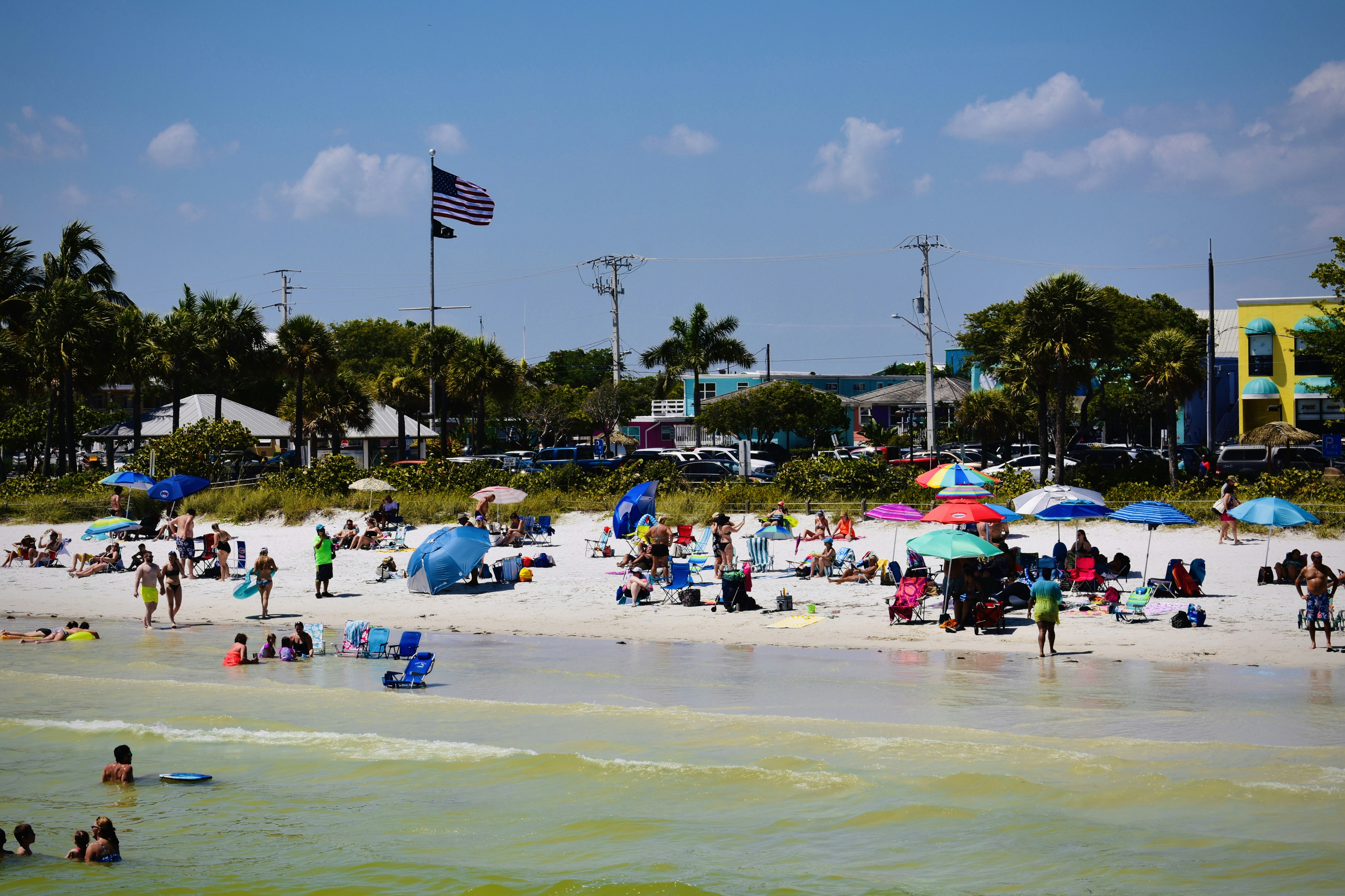 Crowd of beachgoers enjoying a sunny day on the shore, with colorful umbrellas dotting the sandy landscape. American flag waves in the background.