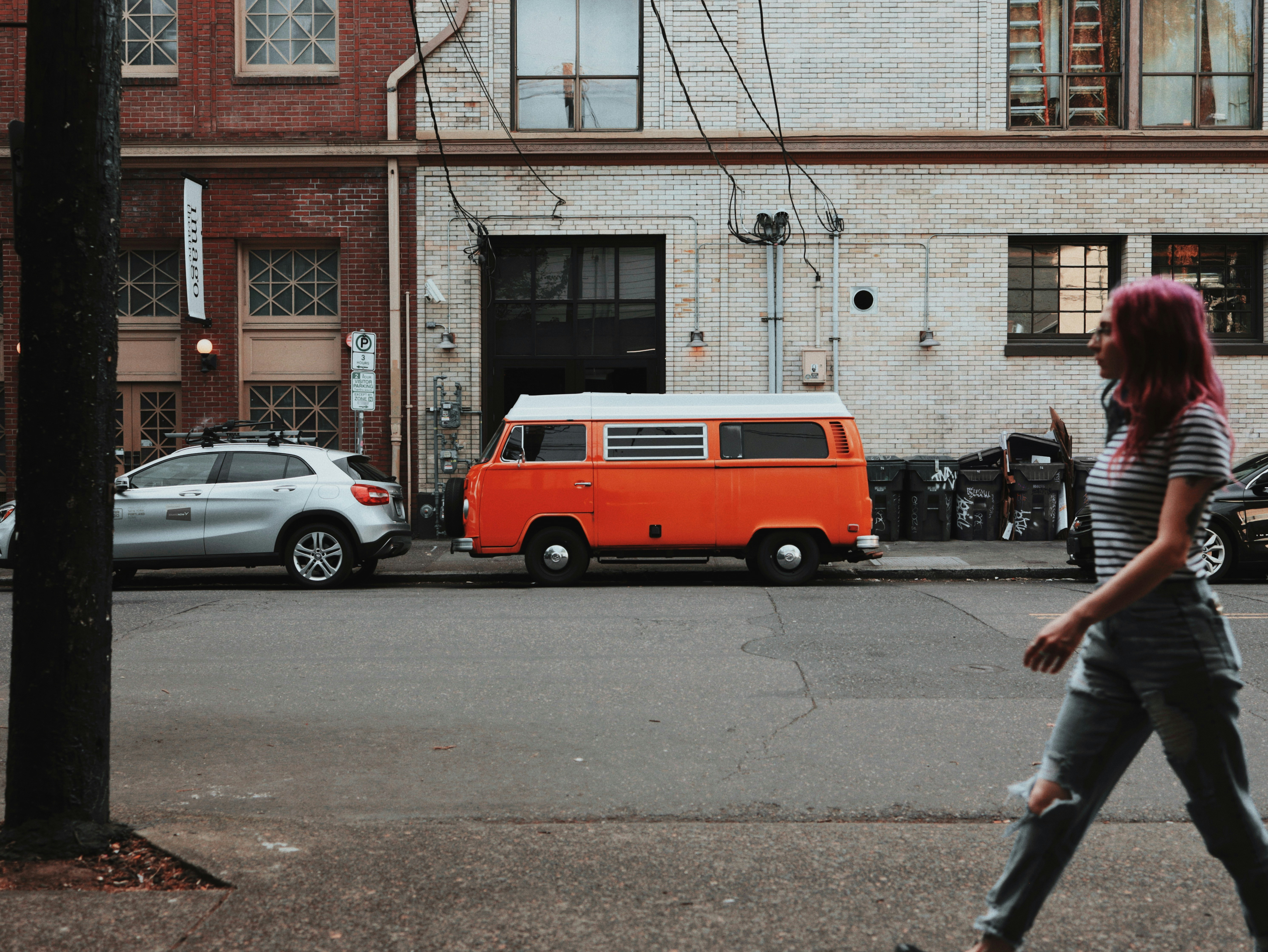 Person with pink hair walking past an orange van parked on a city street.