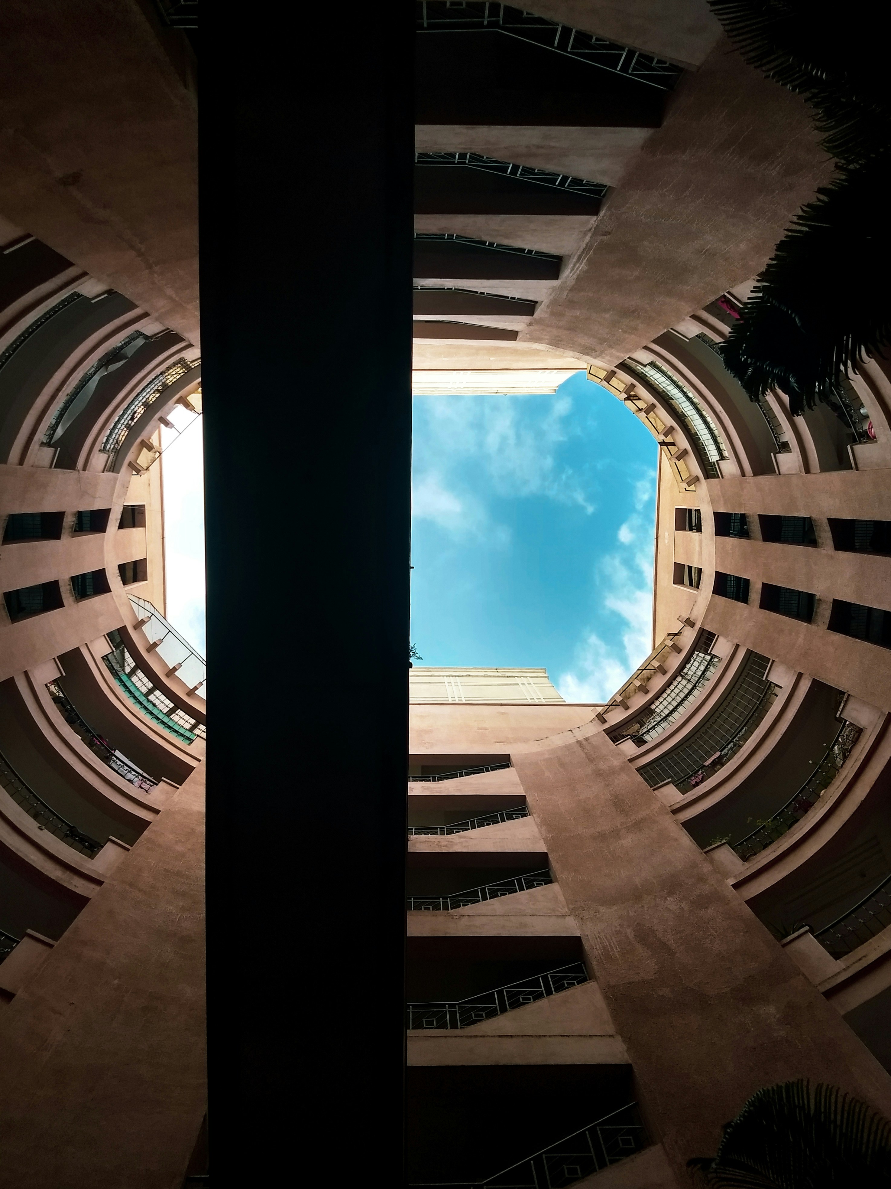 Photograph of a circular, multi-level atrium shot from below, with a dark central column and curved balconies framing a bright blue sky through the opening.